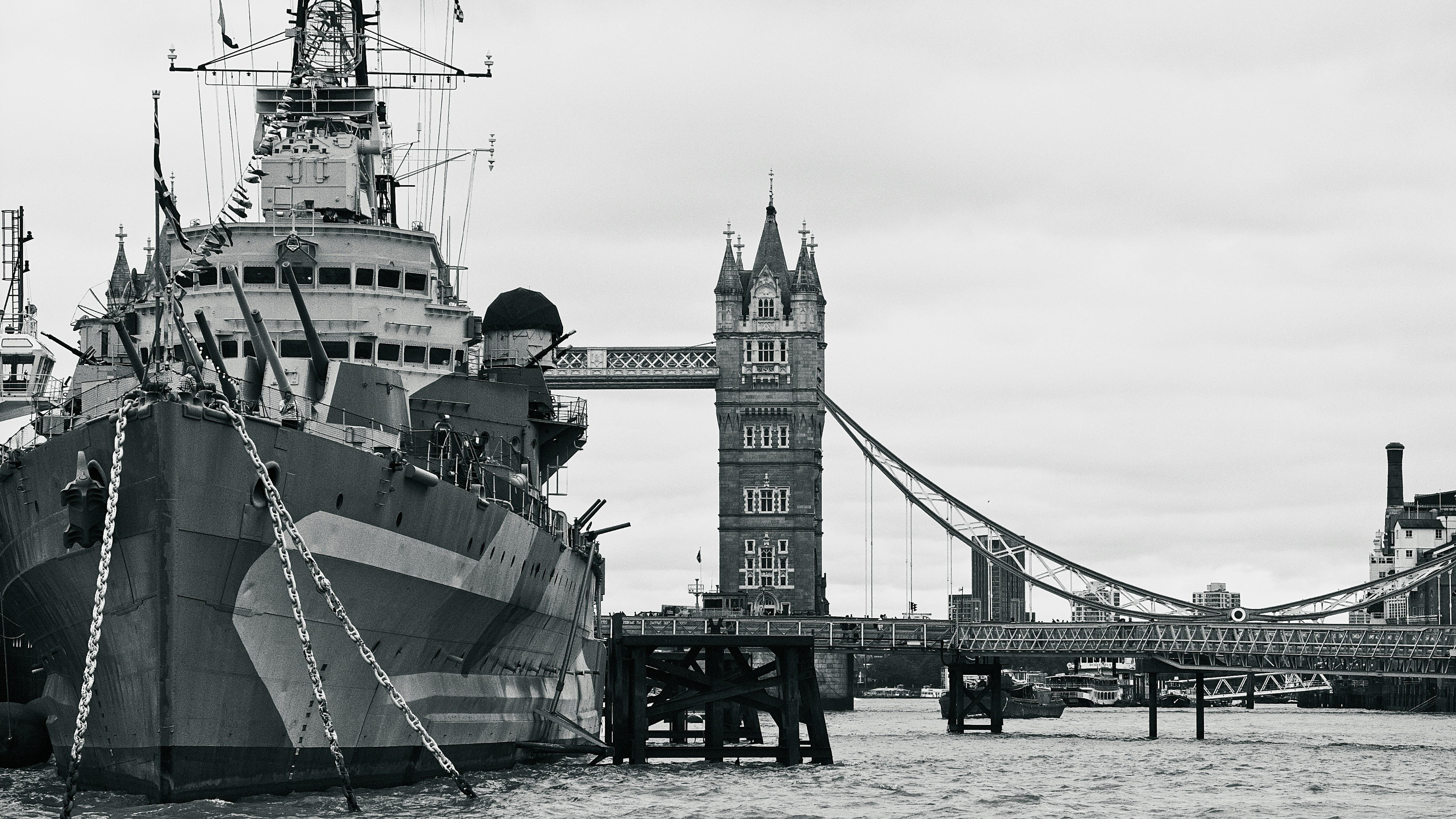 Monochrome photograph of a moored battleship with Tower Bridge rising behind along the Thames.