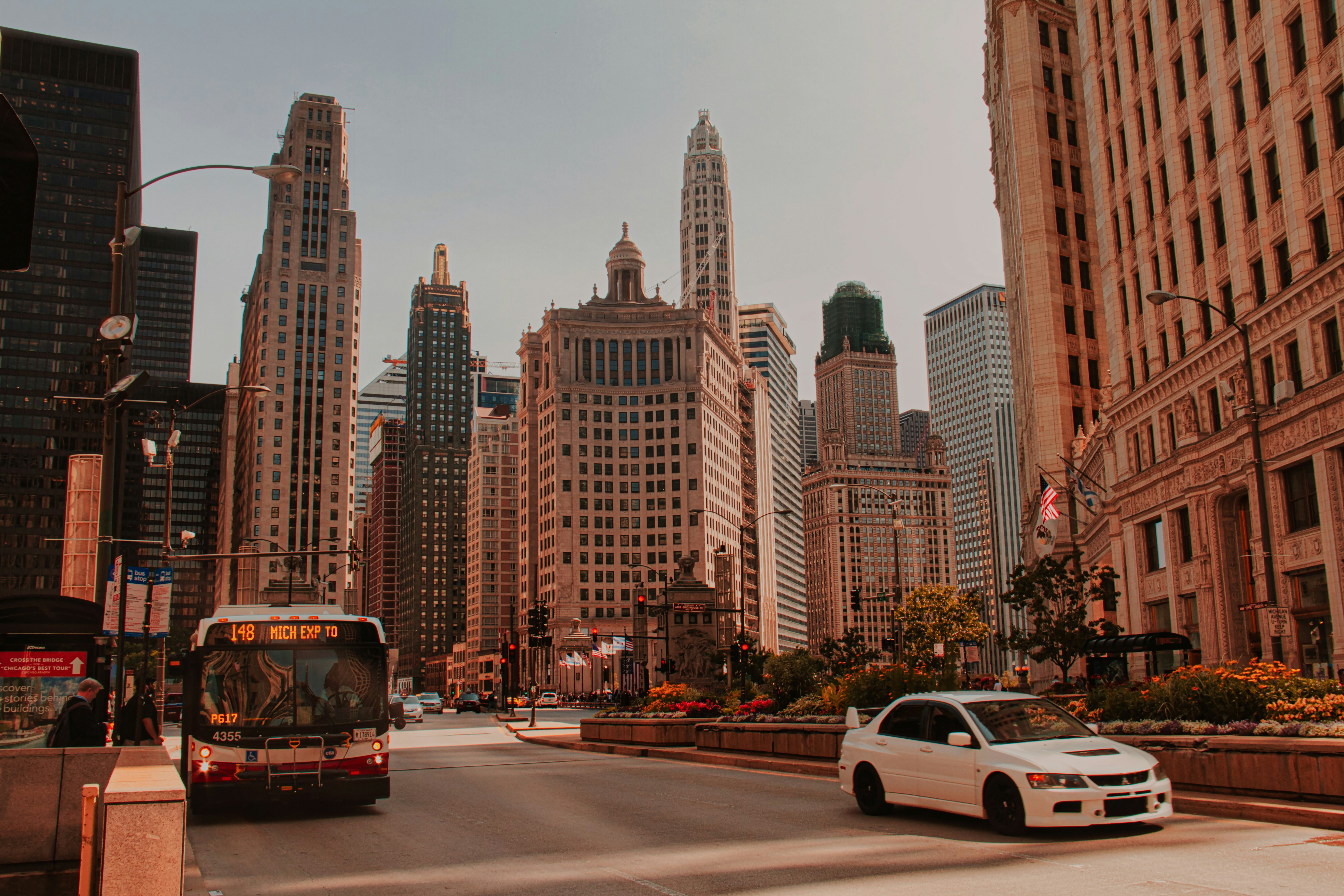 City street bustling with activity, featuring a city bus and a white car amidst towering skyscrapers and vibrant greenery.