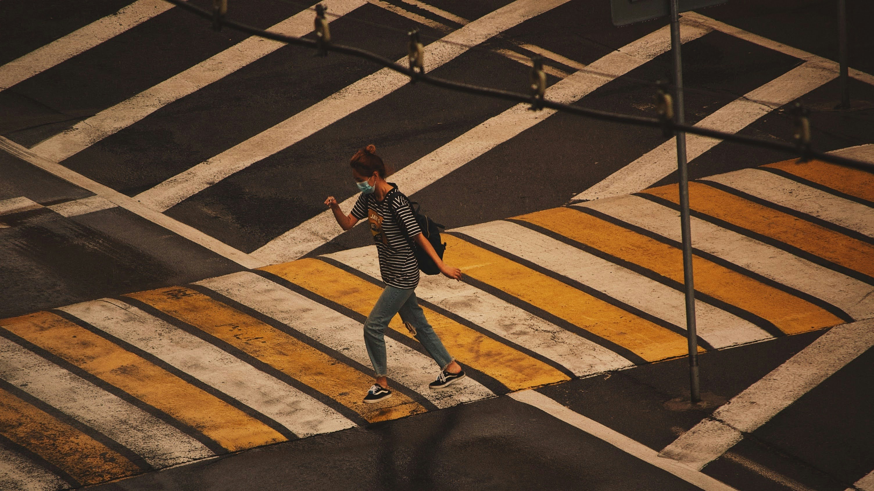 A pedestrian in a striped shirt crosses a yellow and white striped crosswalk in a bustling urban setting. The scene captures the essence of city life.