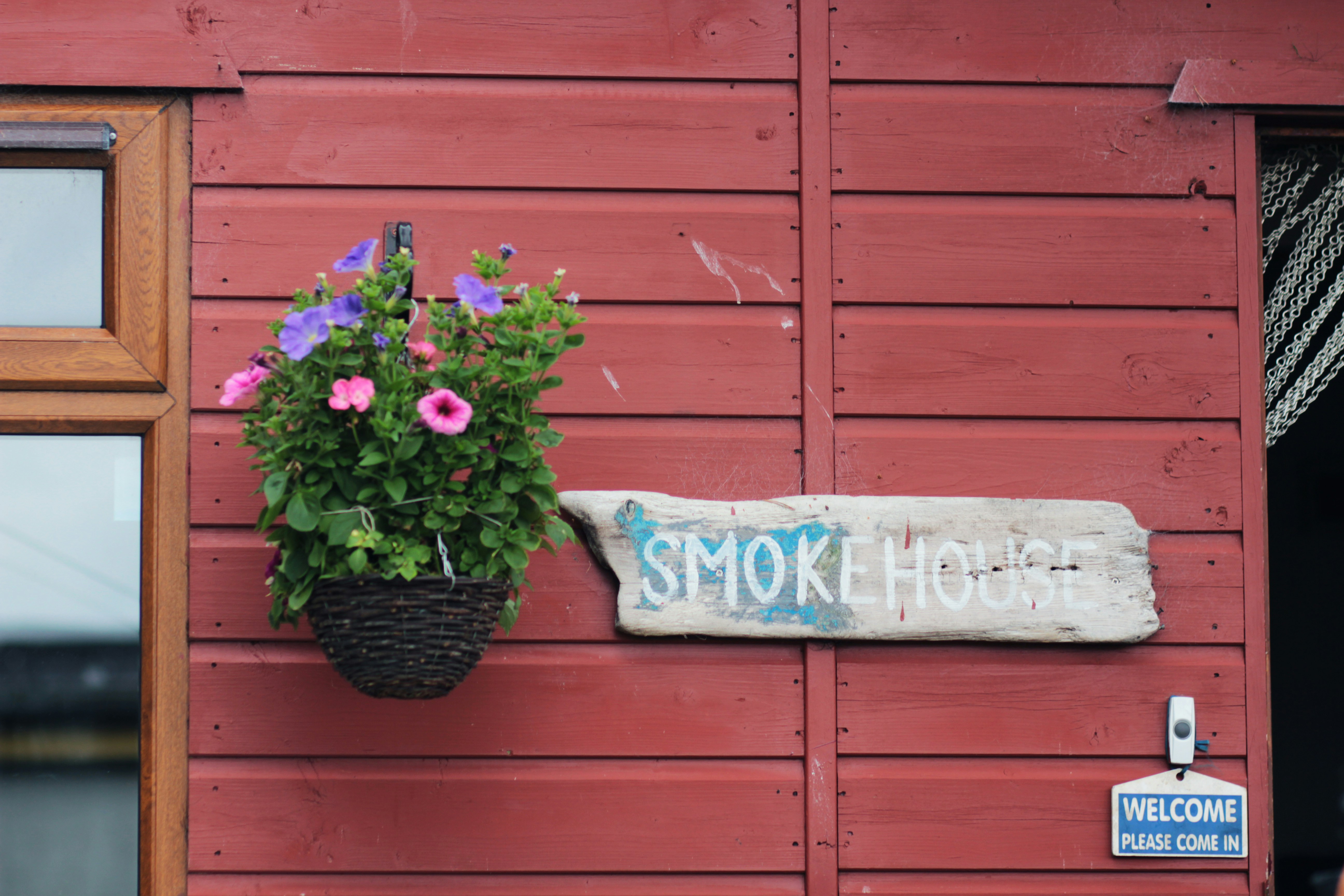 A rustic wooden sign reading 'SMOKE HOUSE' hangs beside a vibrant flower basket on a red wooden wall, inviting visitors in.