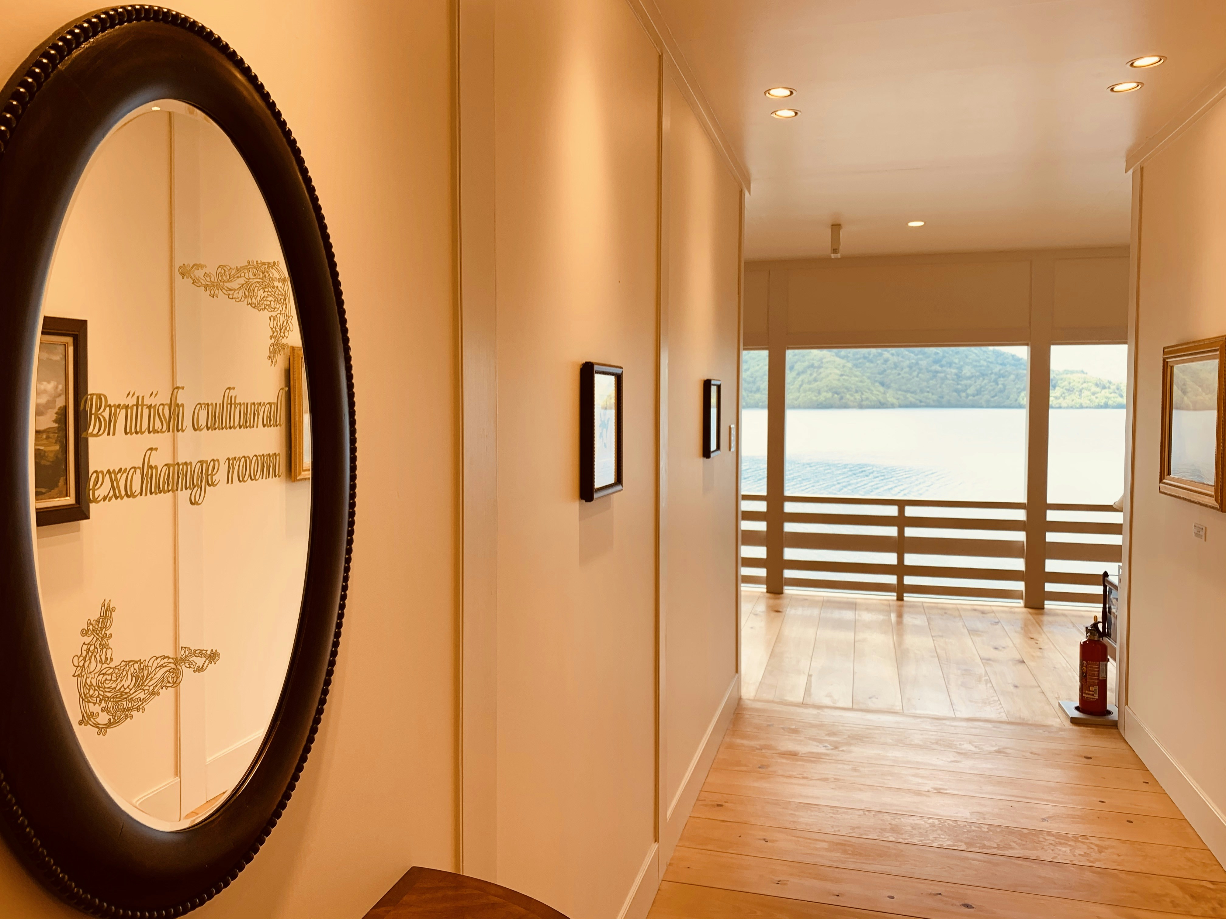 Elegant hallway featuring a round mirror with intricate text, framed artwork, and a view of a tranquil waterfront beyond.