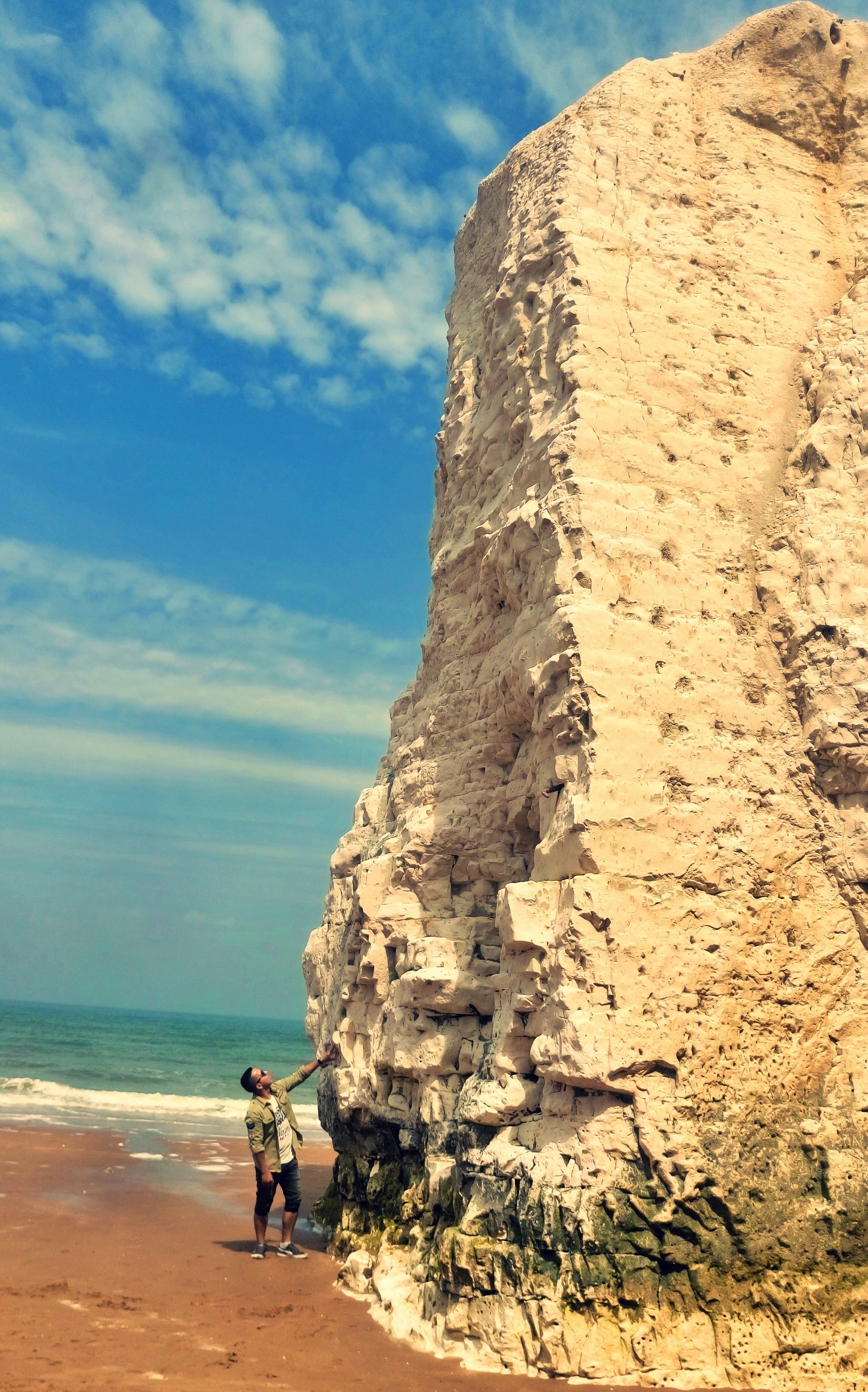 A lone figure stands at the base of a towering white cliff on a sunlit beach, with the ocean and a clear blue sky in the background.