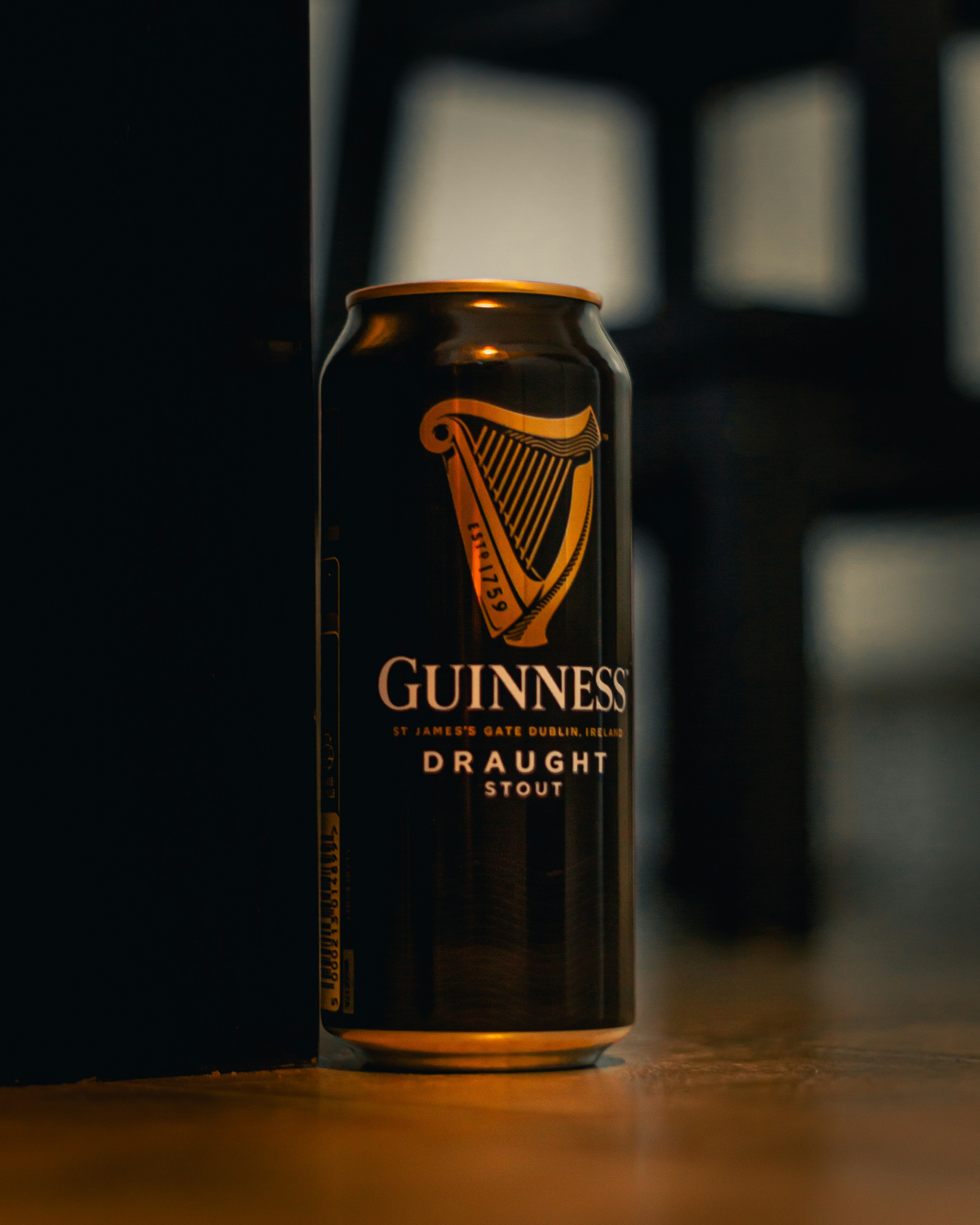 A can of Guinness Draught Stout resting against a dark surface, highlighting its iconic logo and rich colors. The background features a blurred setting, creating a cozy atmosphere.