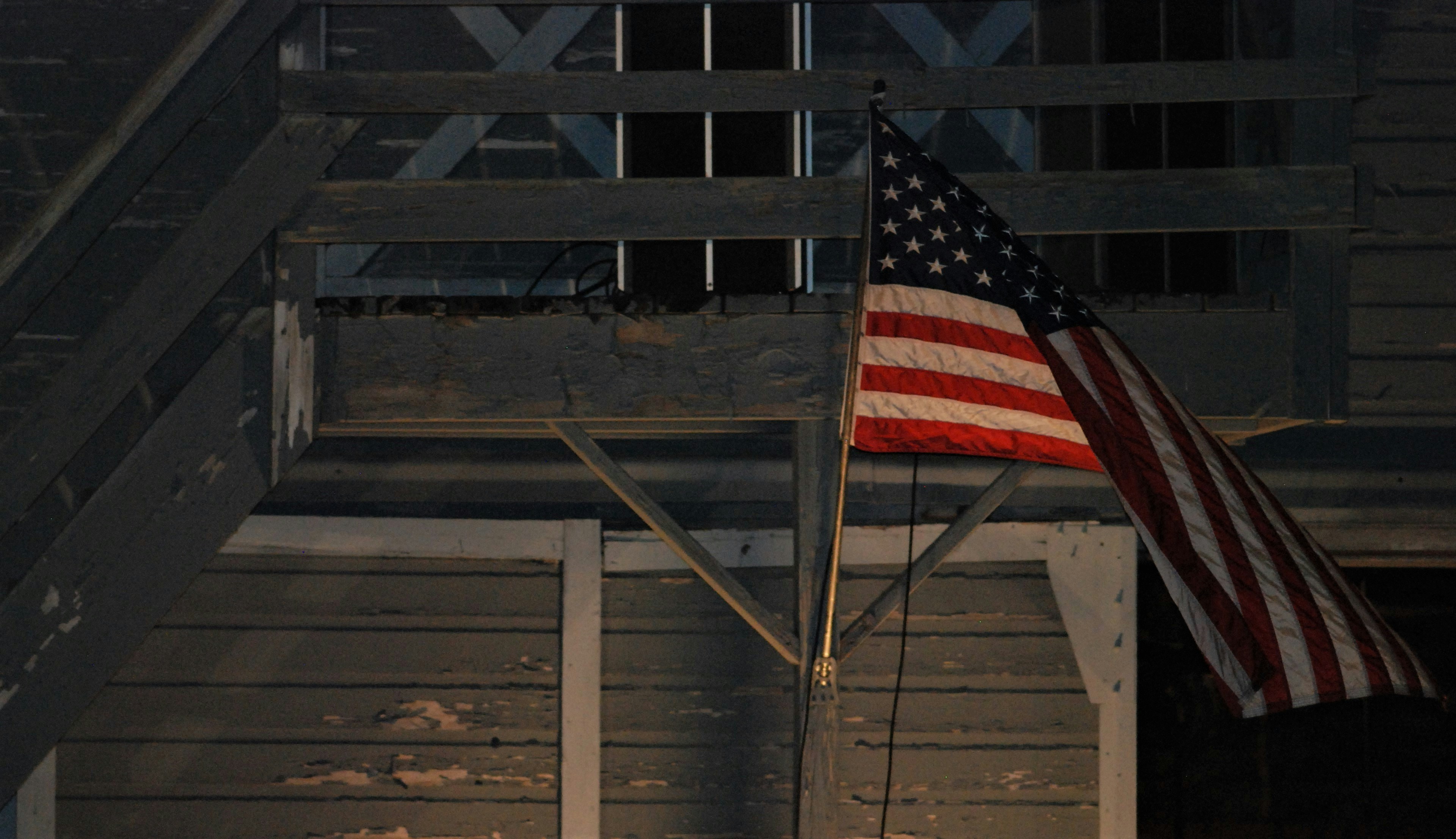 An American flag gently sways against a weathered wooden backdrop, illuminated by soft evening light. The scene evokes a sense of history and nostalgia.