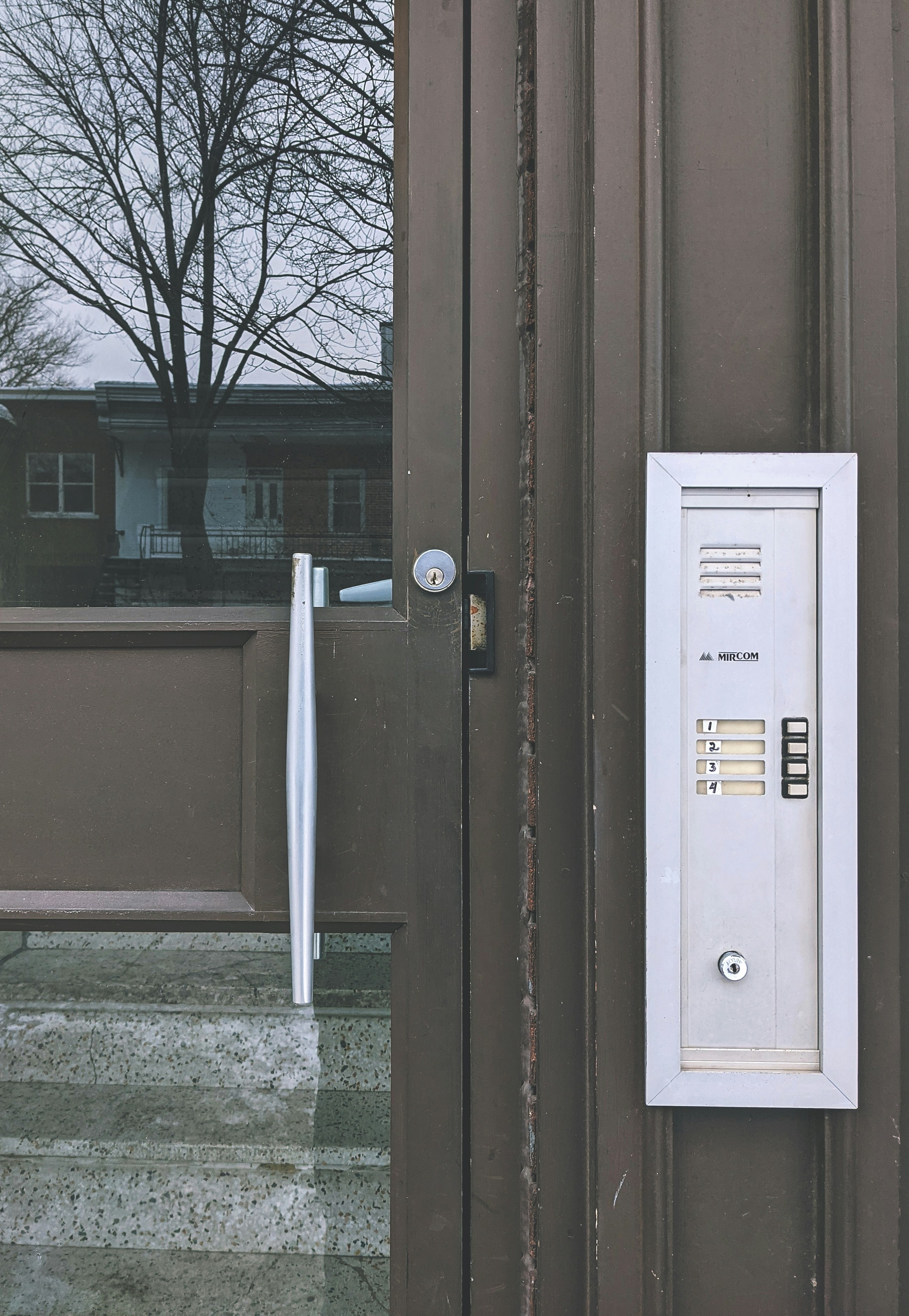 Sleek door handle and intercom system juxtaposed against a reflective glass panel and a bare tree in the background.