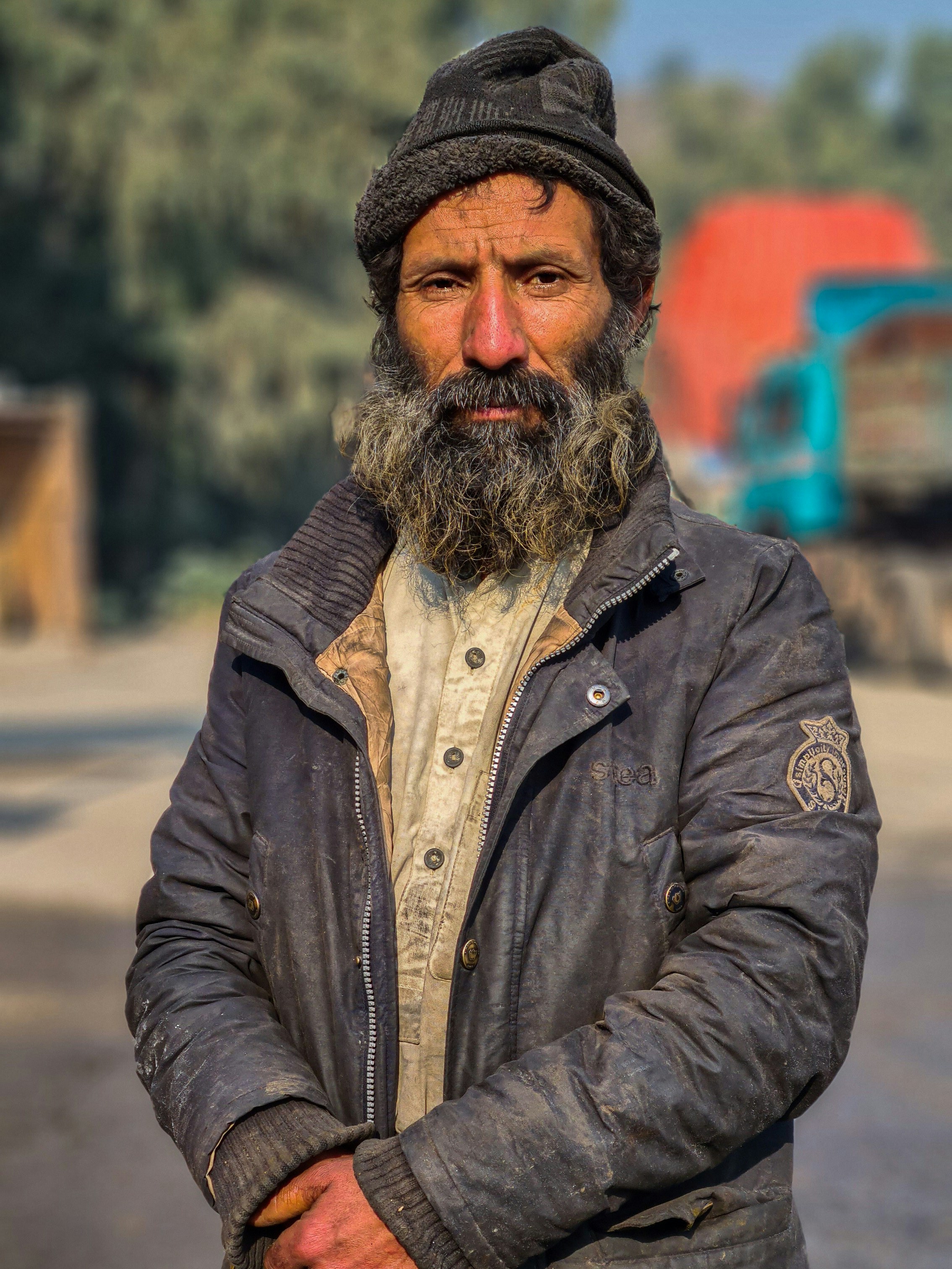 A rugged man with a thick beard and a worn jacket stands with his arms crossed, exuding a sense of strength and endurance against a blurred backdrop of vehicles.
