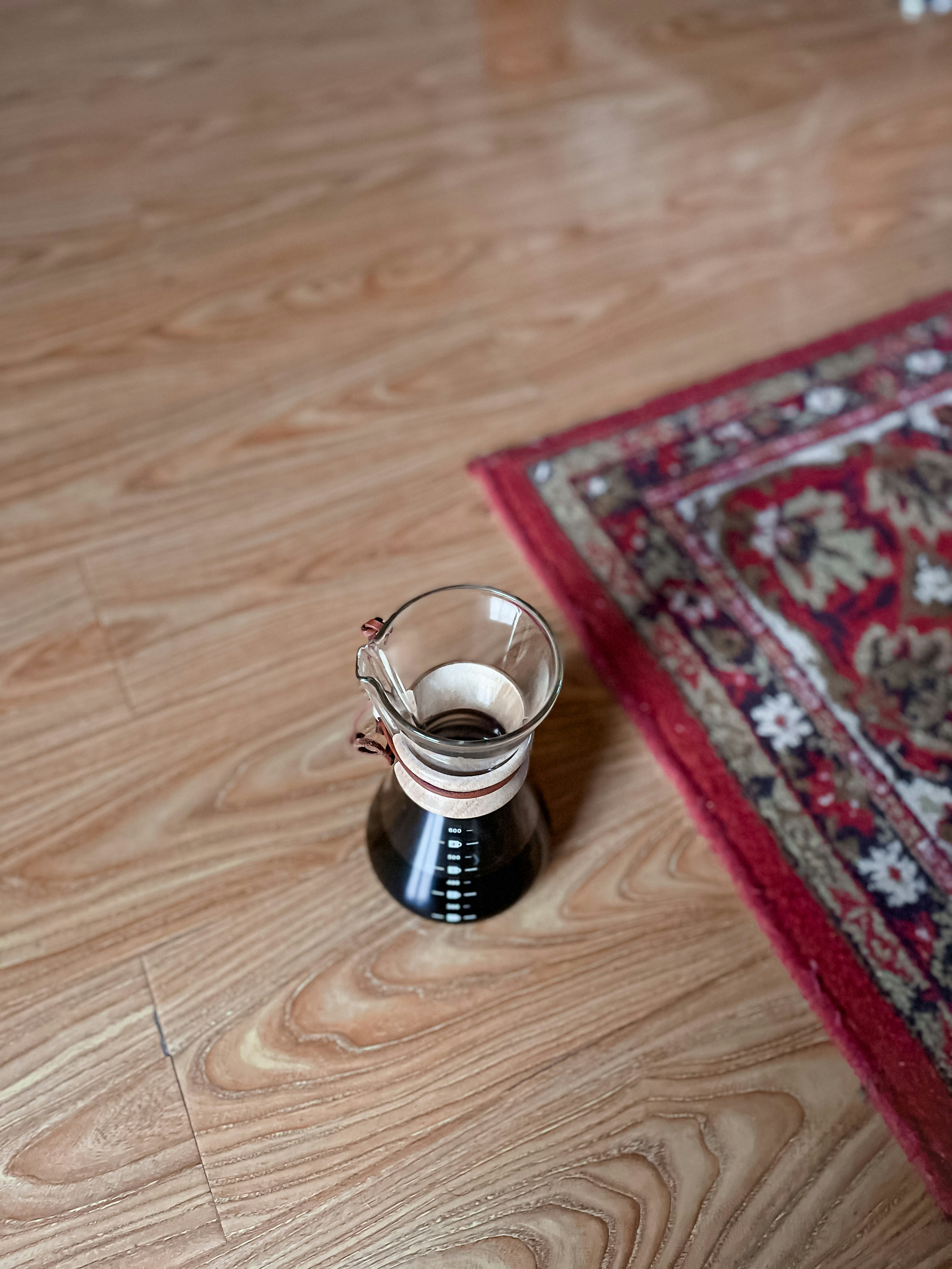 Glass Pour-Over Dripper Resting on a Sunlit Wooden Floor Beside a Red Rug