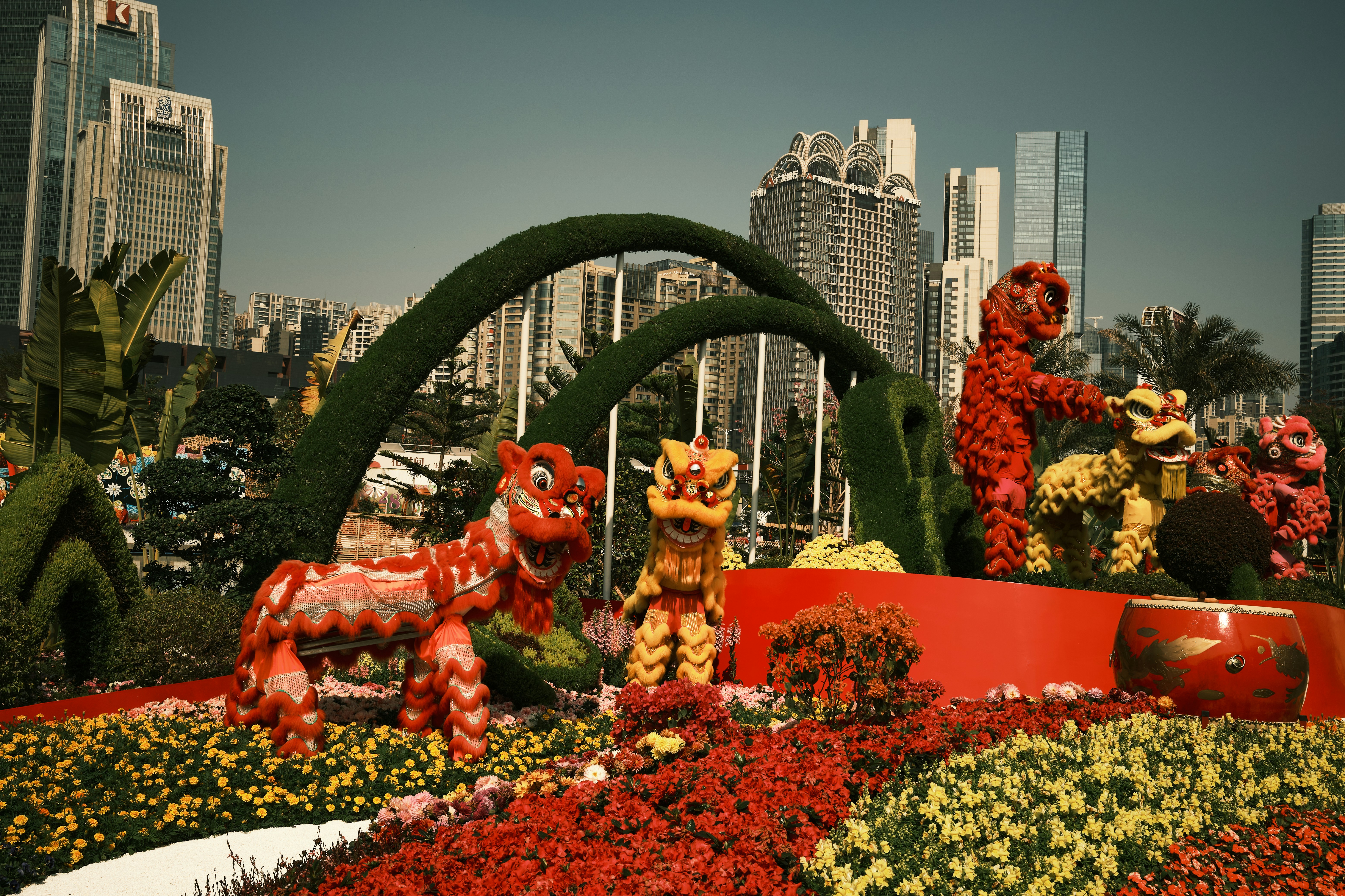 Vibrant floral sculptures of mythical creatures amidst a backdrop of towering skyscrapers. The scene showcases a blend of nature and urban architecture.