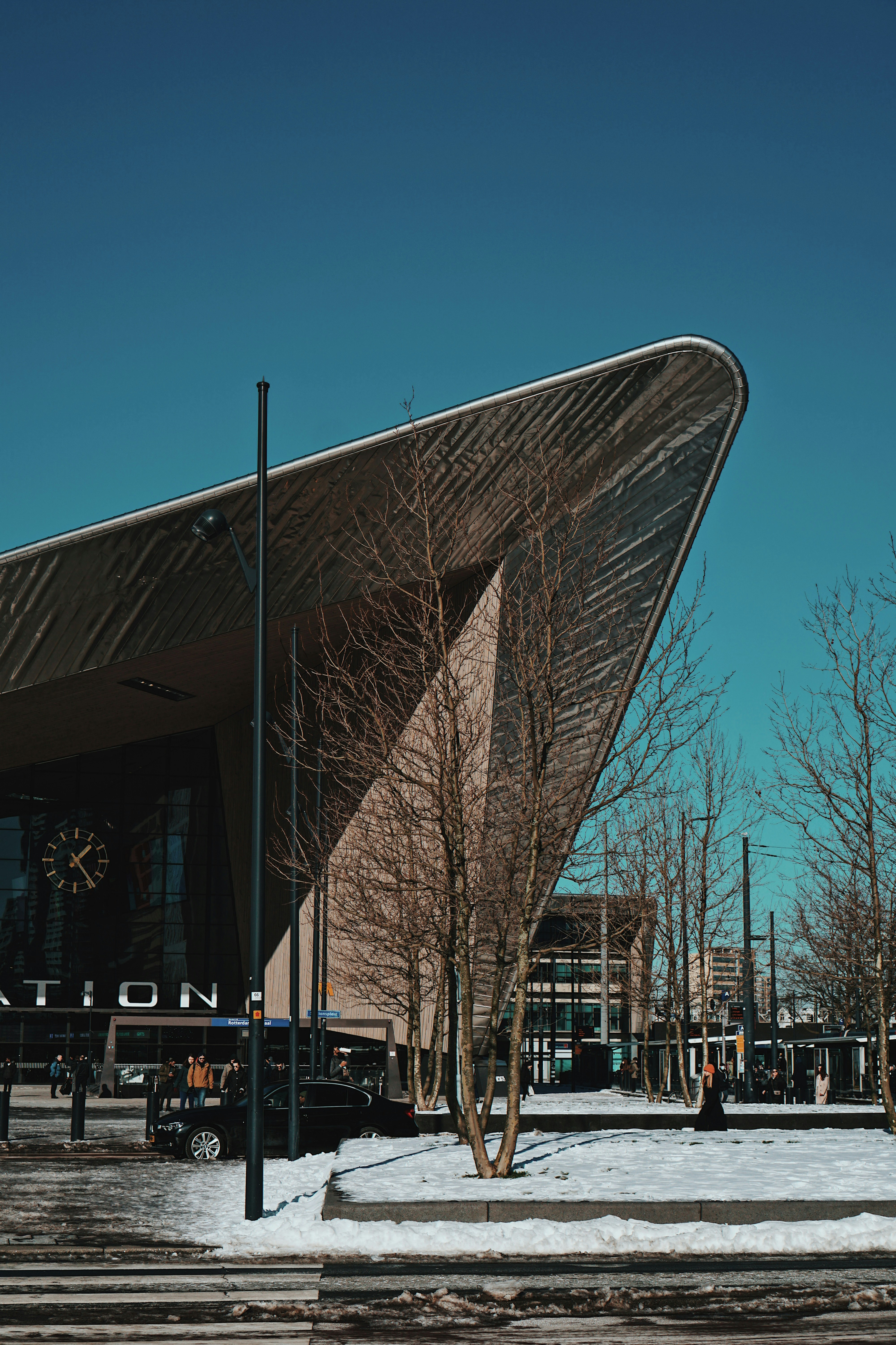 Modern train station with a striking angular roof, surrounded by leafless trees and a snowy landscape. The scene captures the blend of architecture and daily urban activities.