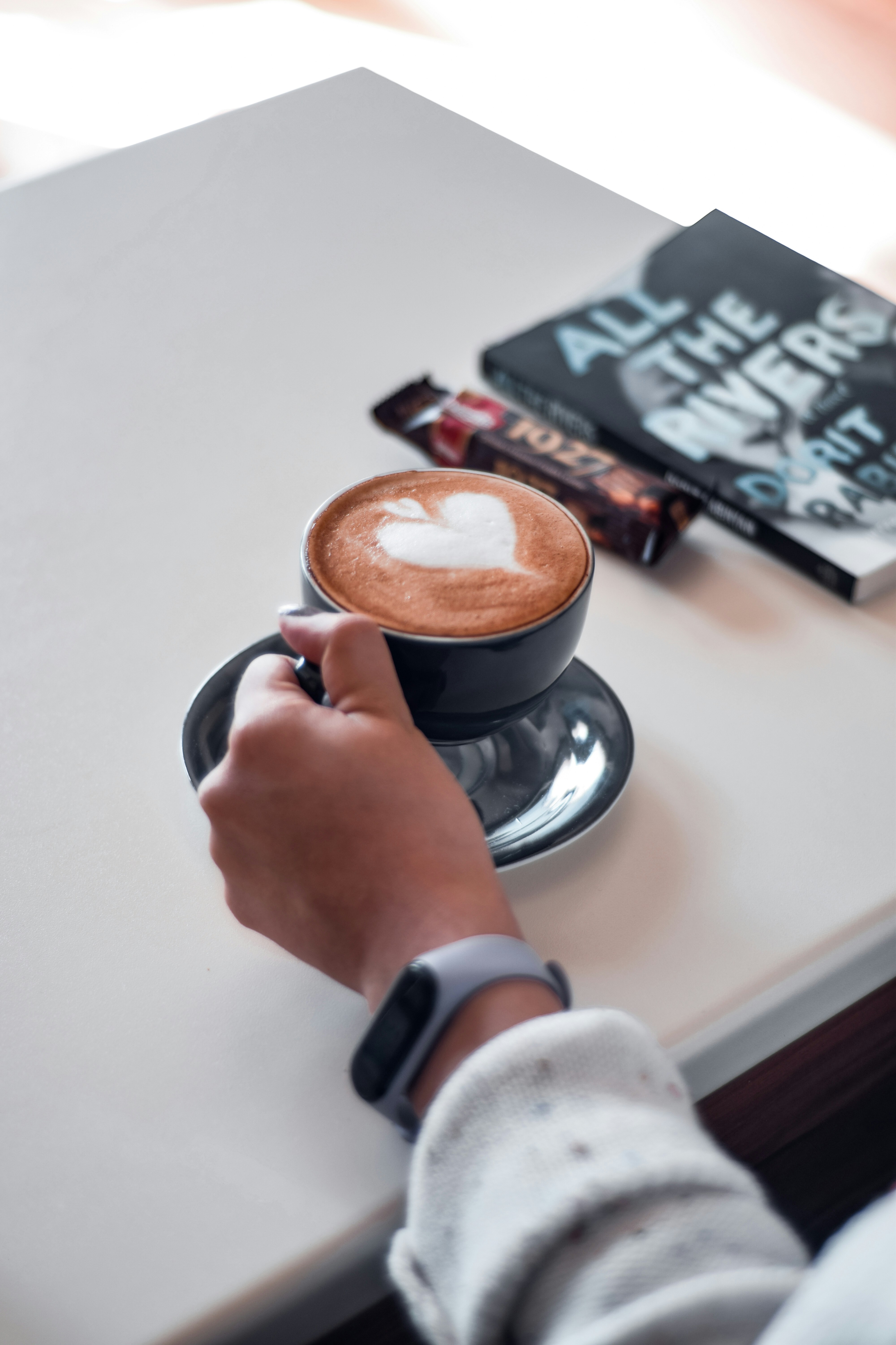 Hand holding a coffee cup with heart-shaped latte art, accompanied by a chocolate bar and a book on a table.