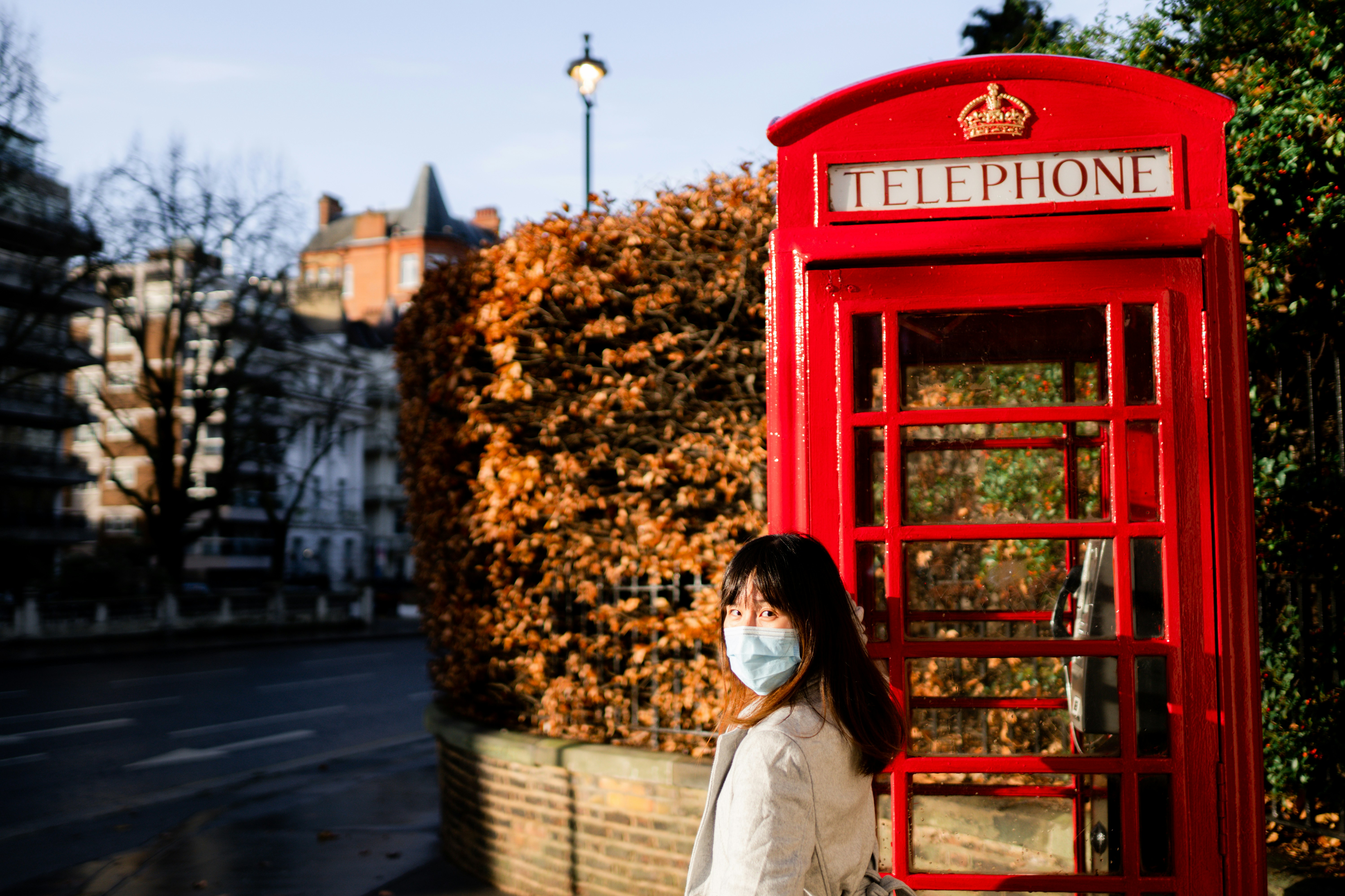 A woman wearing a mask stands beside a classic red telephone booth, surrounded by autumn foliage in a cityscape.
