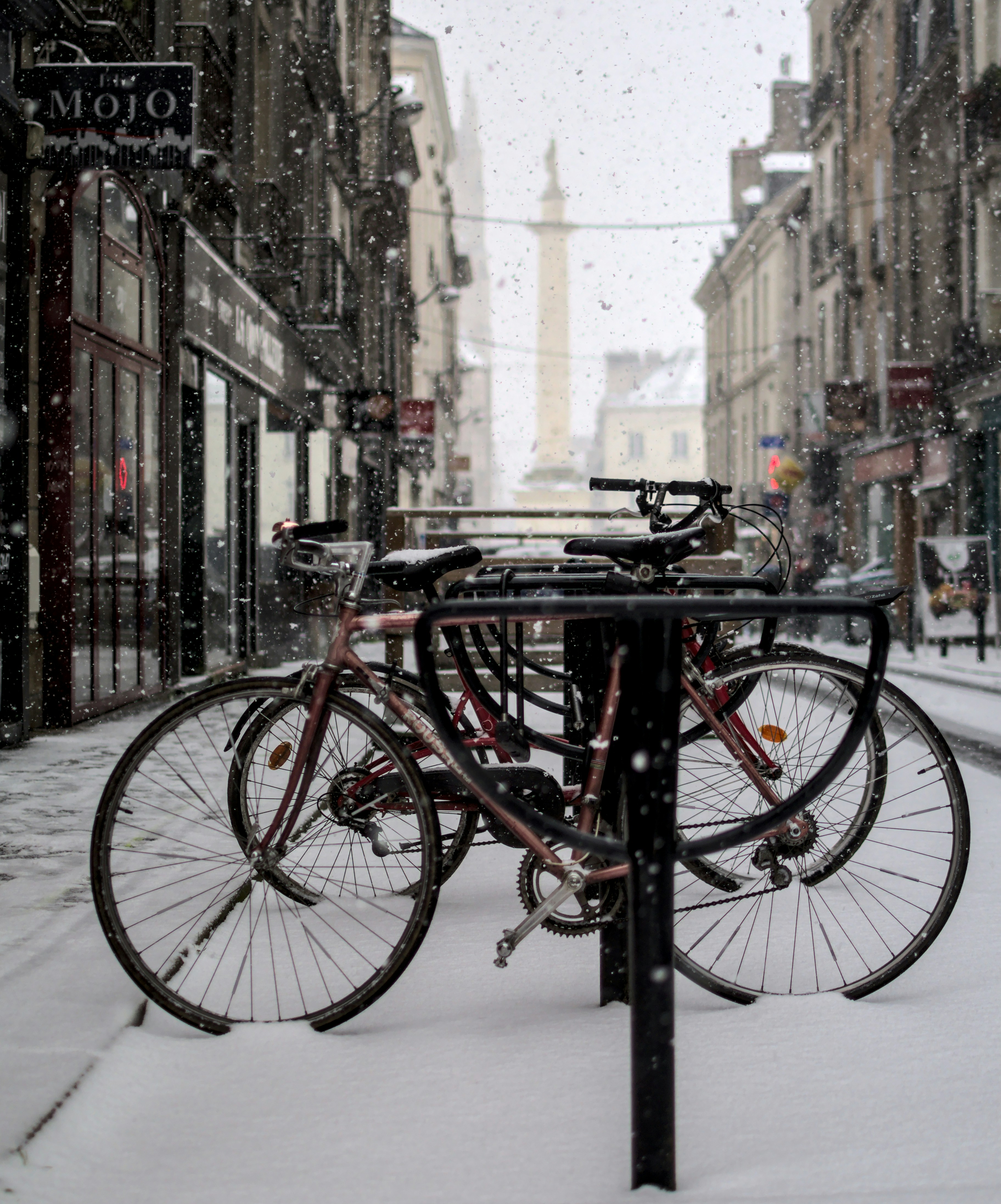 Bicycles parked in a snow-covered street, with a monument visible in the distance. Snowflakes gently fall, creating a serene atmosphere.