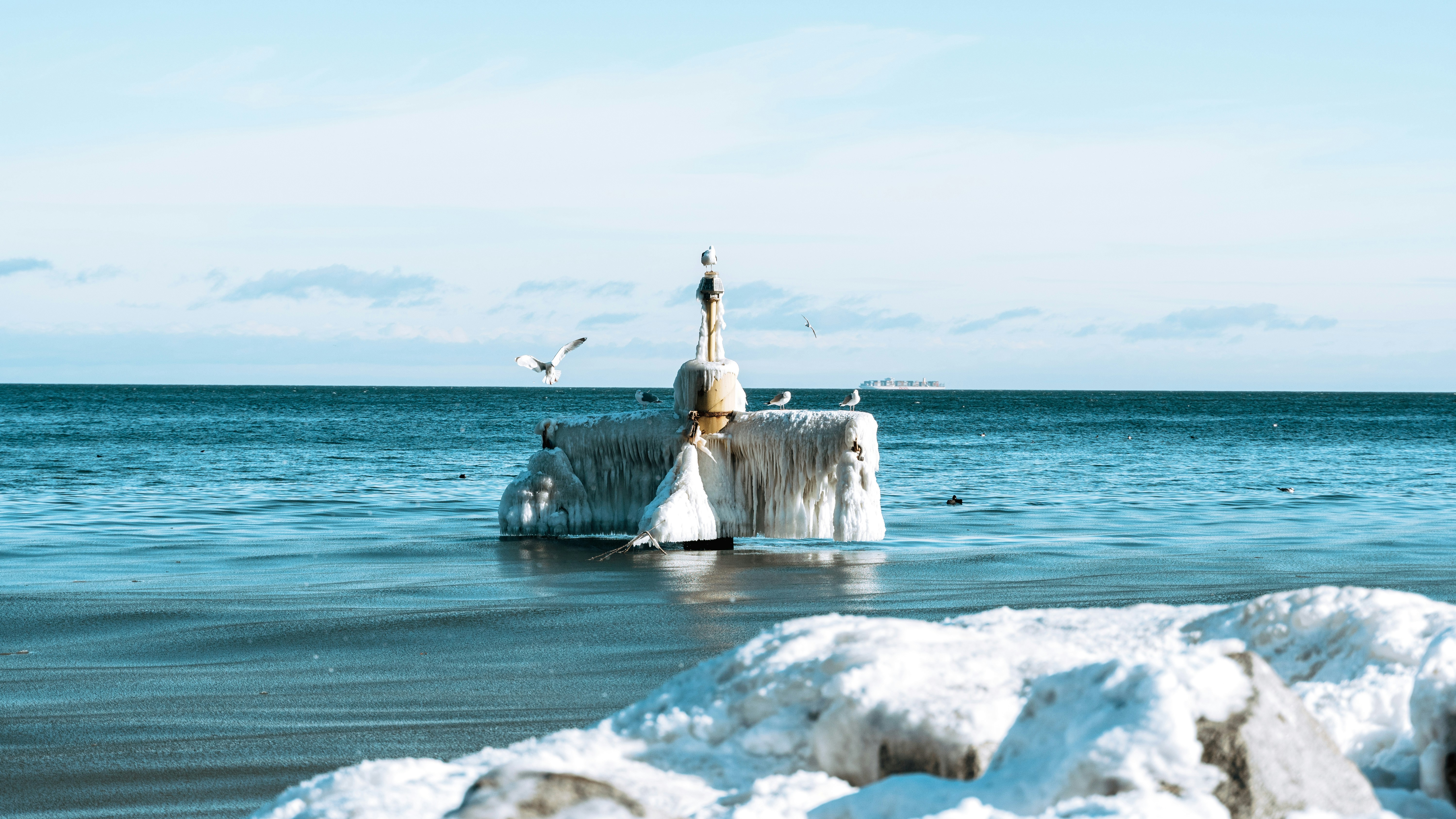 Ice-covered lighthouse standing in a tranquil sea under a clear blue sky.