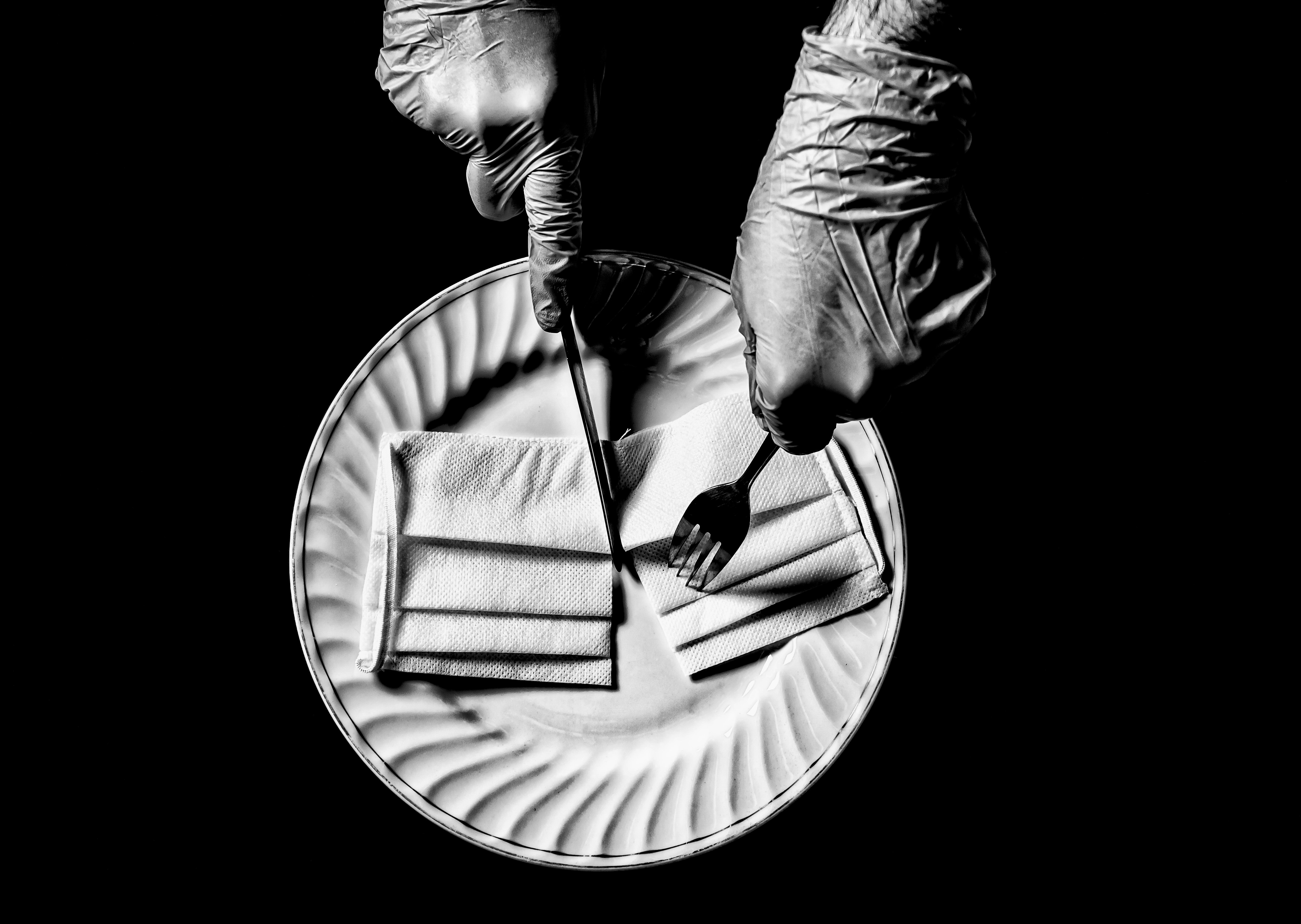 Gloved hands cutting a stack of paper squares with a fork and knife on a paper plate.
