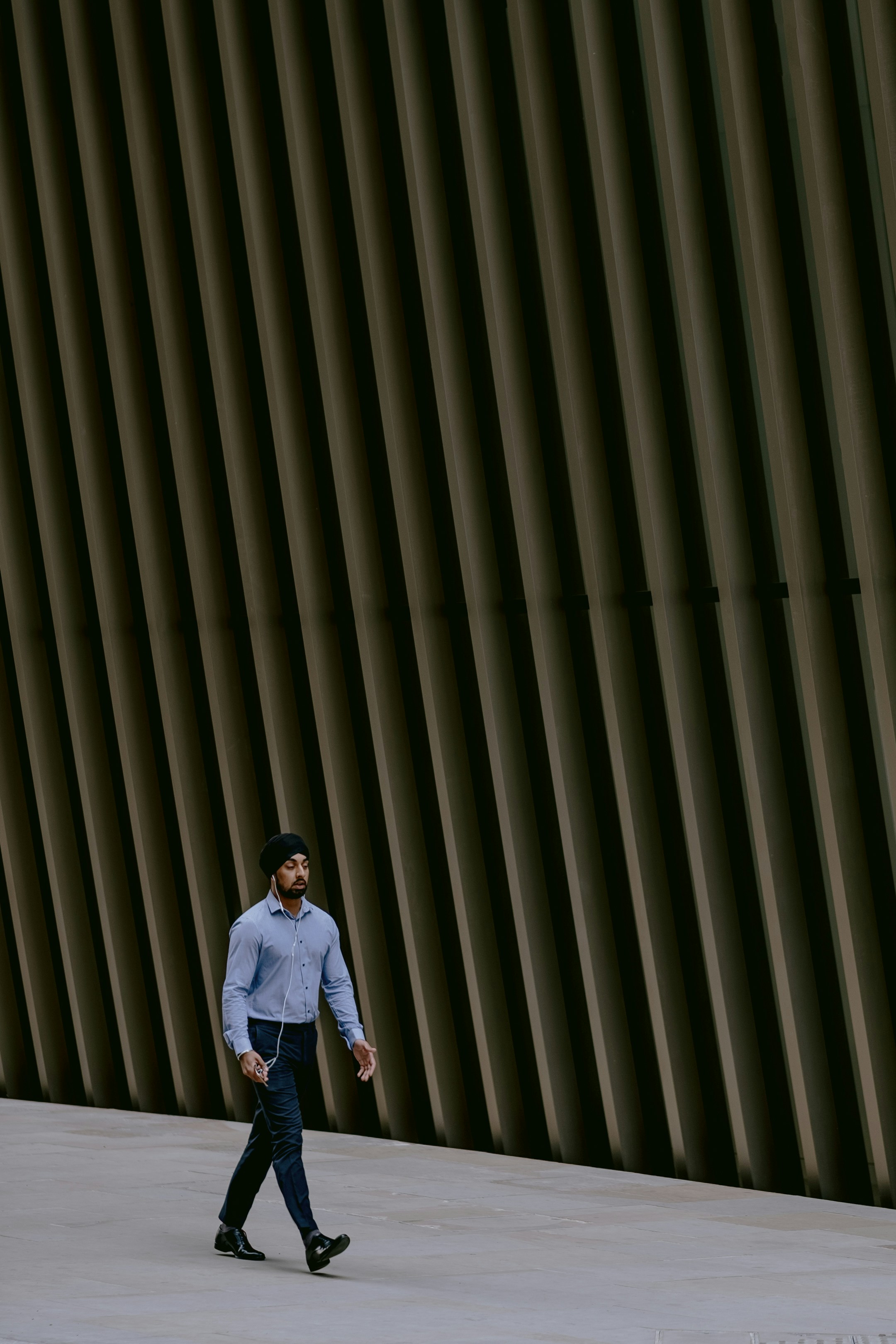 A man in a blue shirt and dark pants walks along a textured wall with vertical lines, embodying a blend of fashion and architectural design.