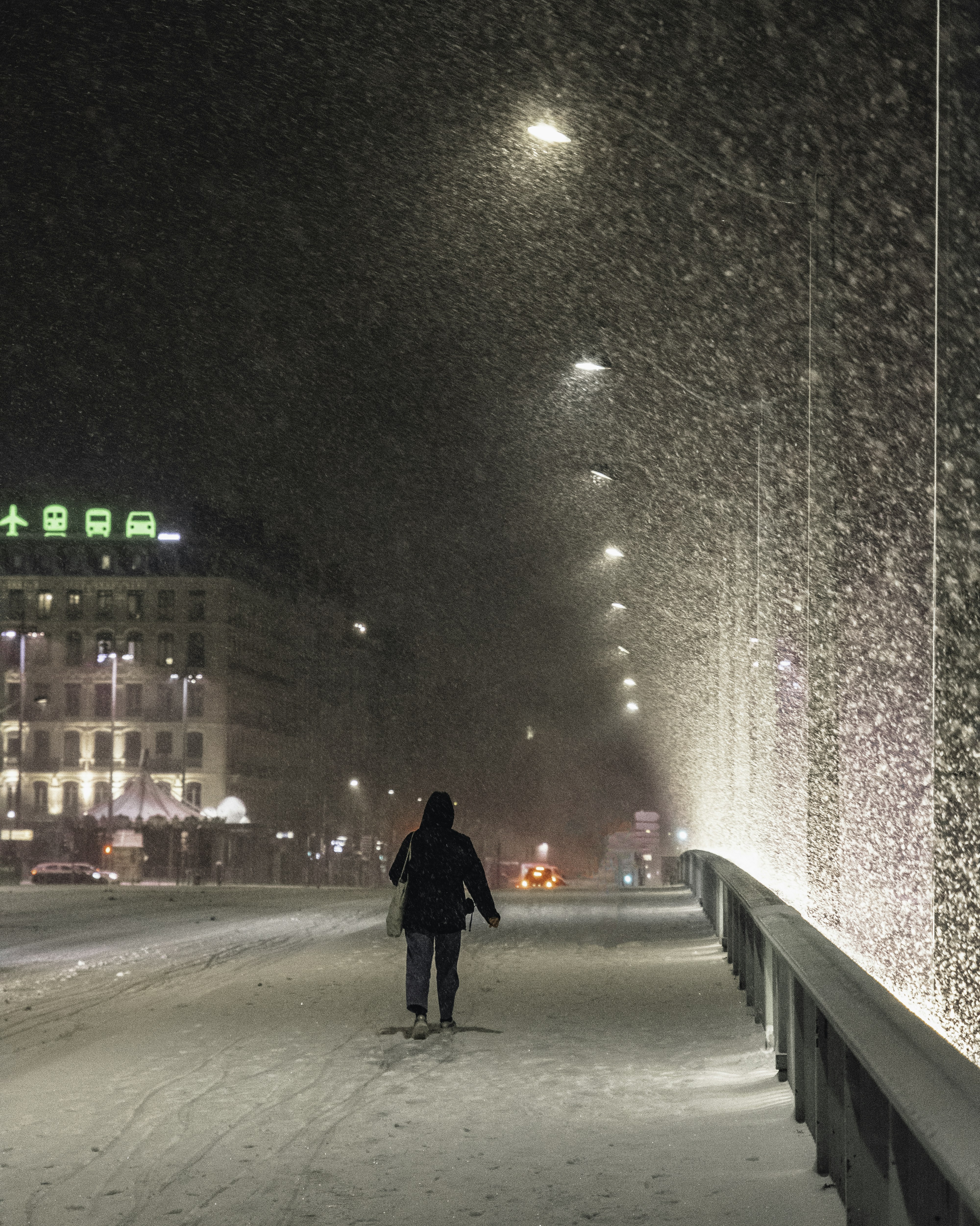 A solitary figure walks through a heavy snowfall along a dimly lit street, illuminated by streetlights and distant buildings. The scene conveys a sense of solitude amidst the winter storm.