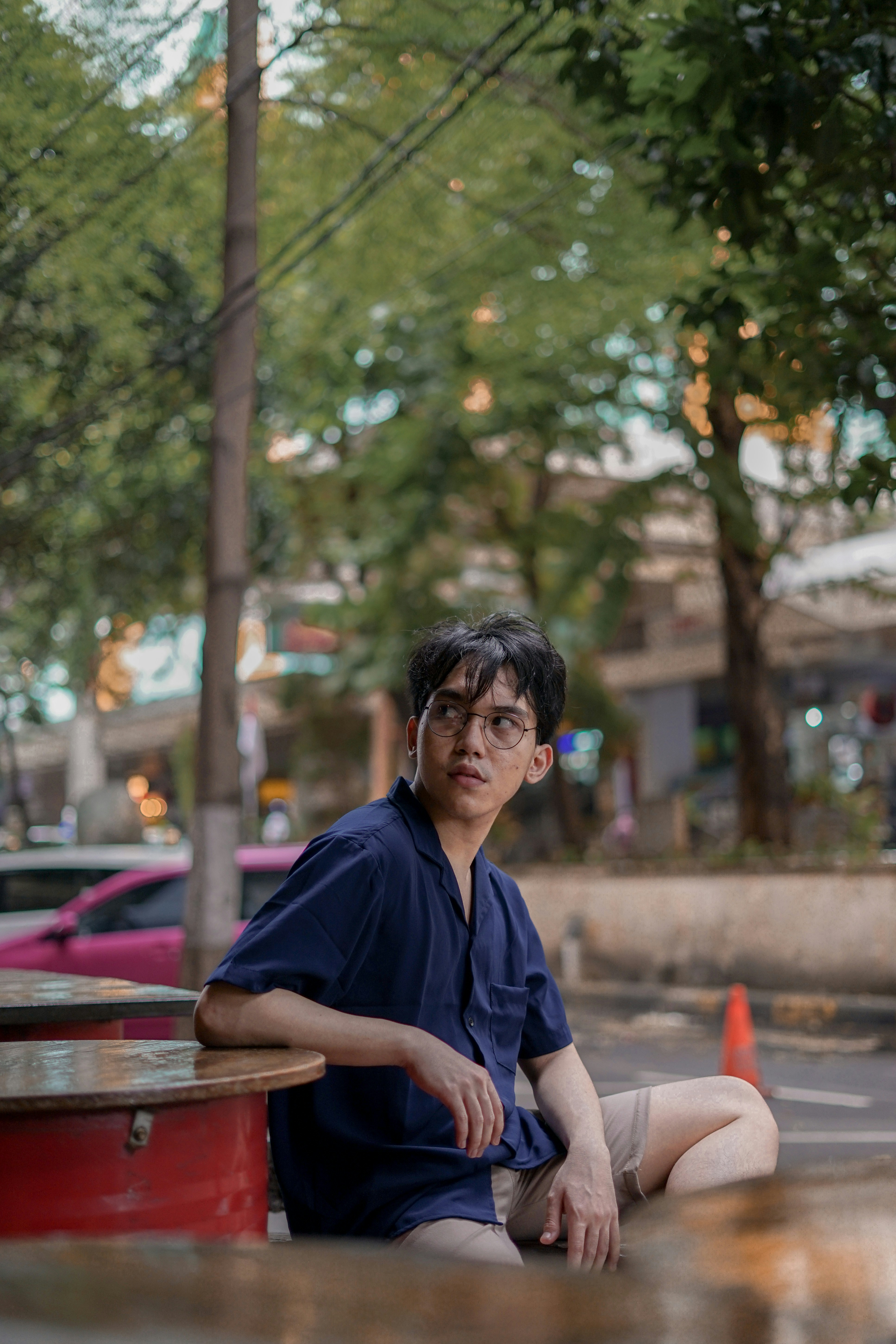 Young man seated on a ledge, gazing thoughtfully amidst a vibrant urban backdrop with blurred street elements. 
