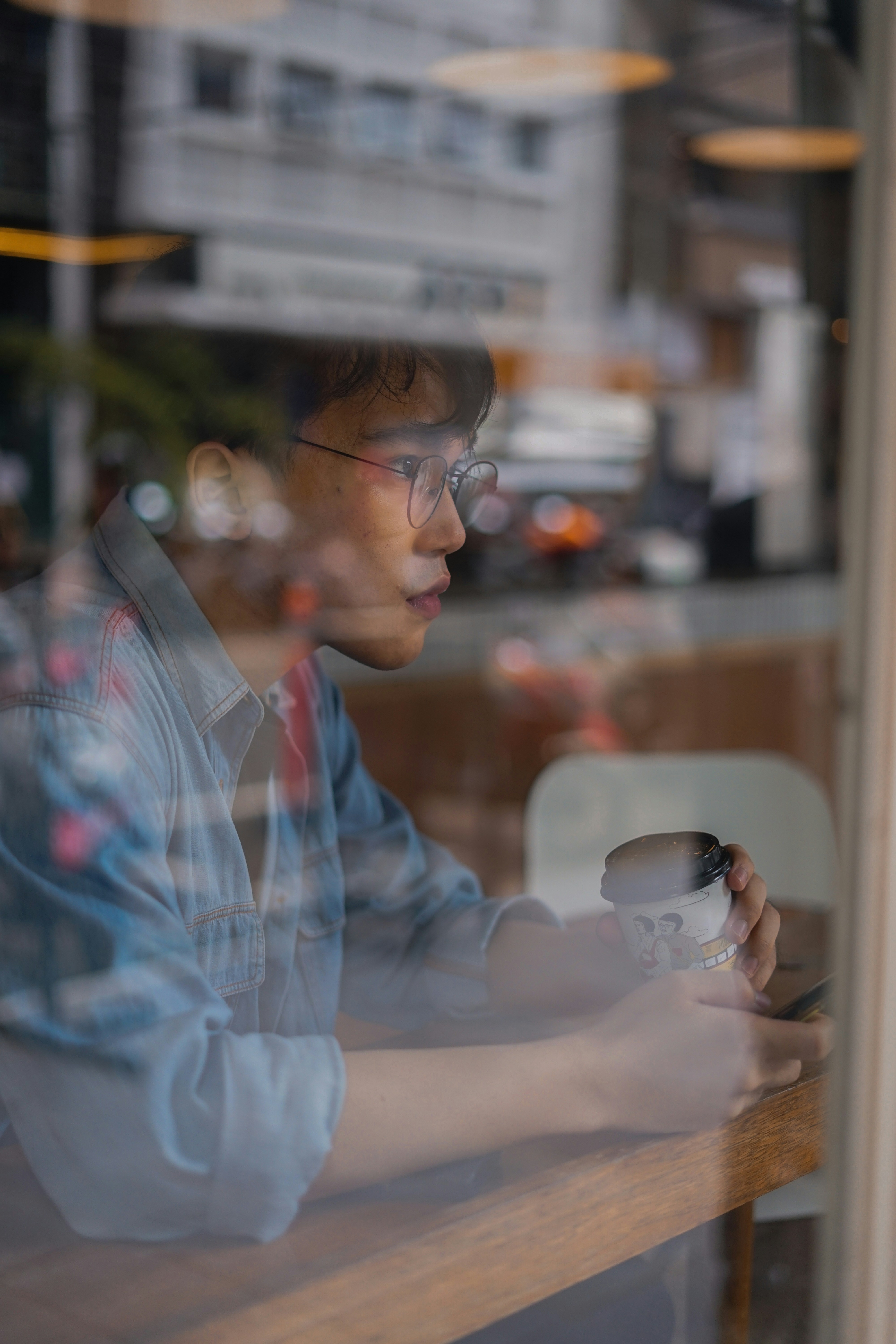Young man sitting at a café table, holding a cup while gazing thoughtfully through a window. The scene captures urban life with reflections adding depth.