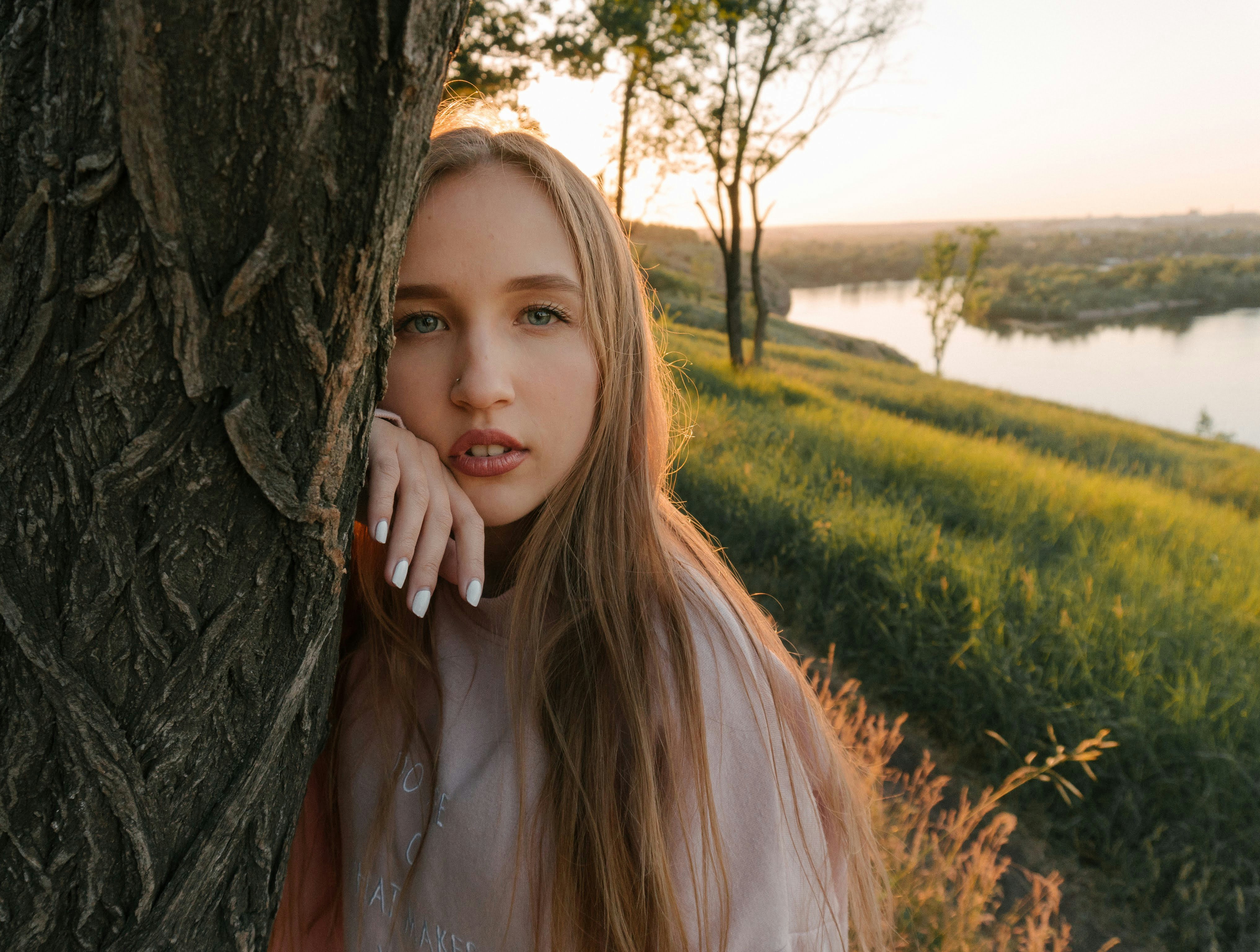 A young woman leans against a tree, her gaze contemplative as the sun sets over a tranquil river landscape.