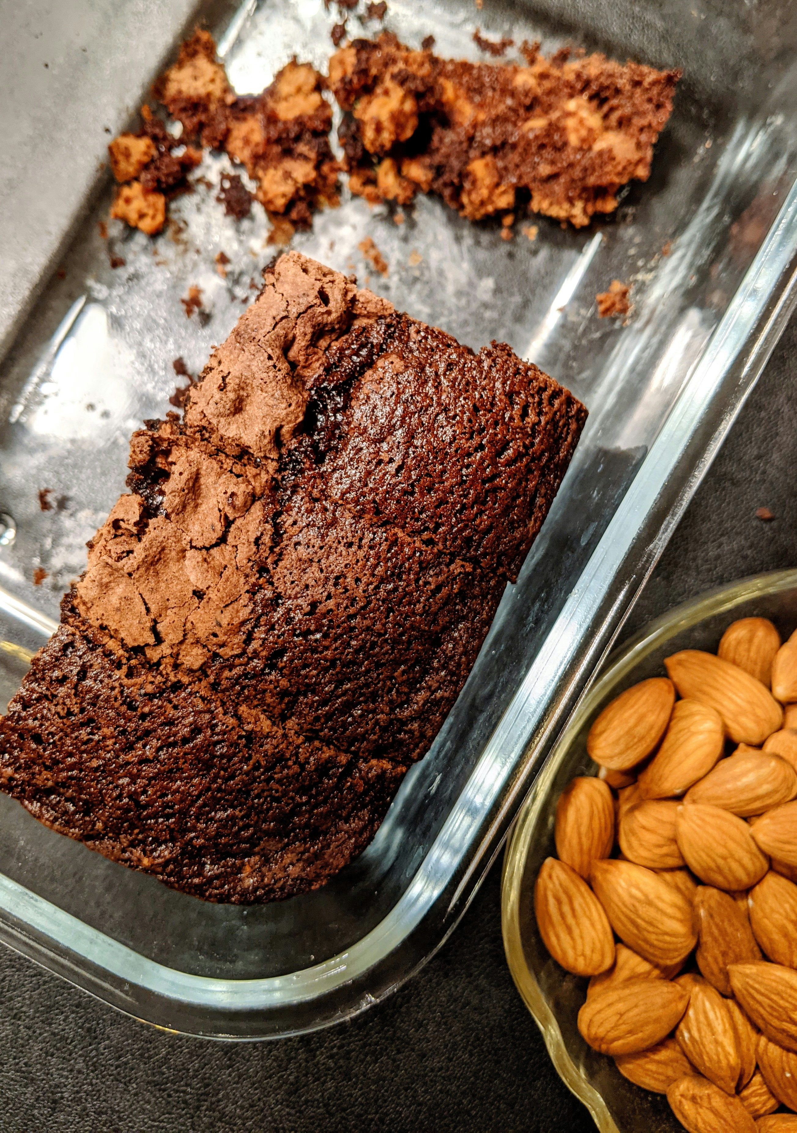 Rich chocolate brownie resting in a glass dish, accompanied by a bowl of almonds. Crumbled pieces hint at indulgent flavors.