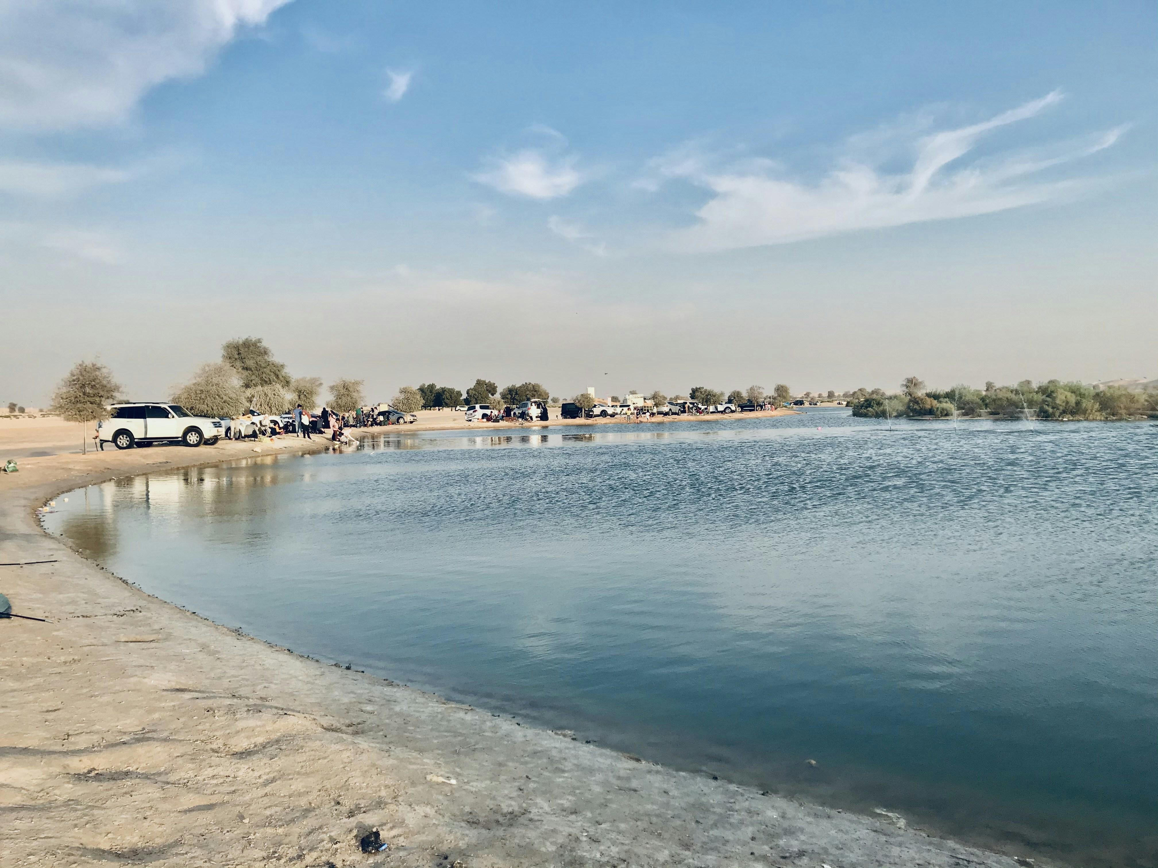 Calm waters reflect the sky, bordered by a sandy shore and distant figures enjoying the landscape.