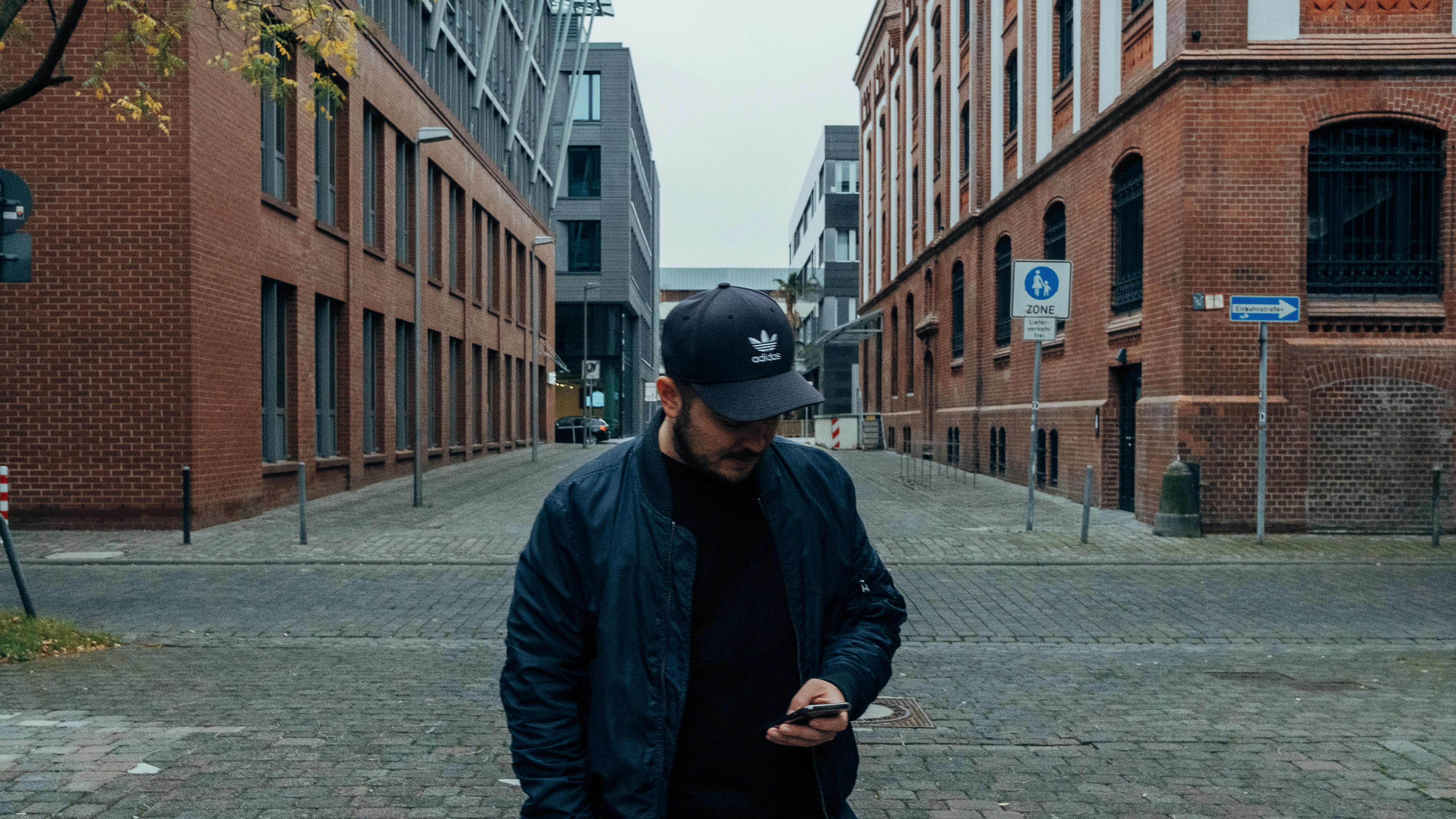 A man in a black jacket and cap stands in a cobblestone street, absorbed in his phone, surrounded by modern and historic architecture.