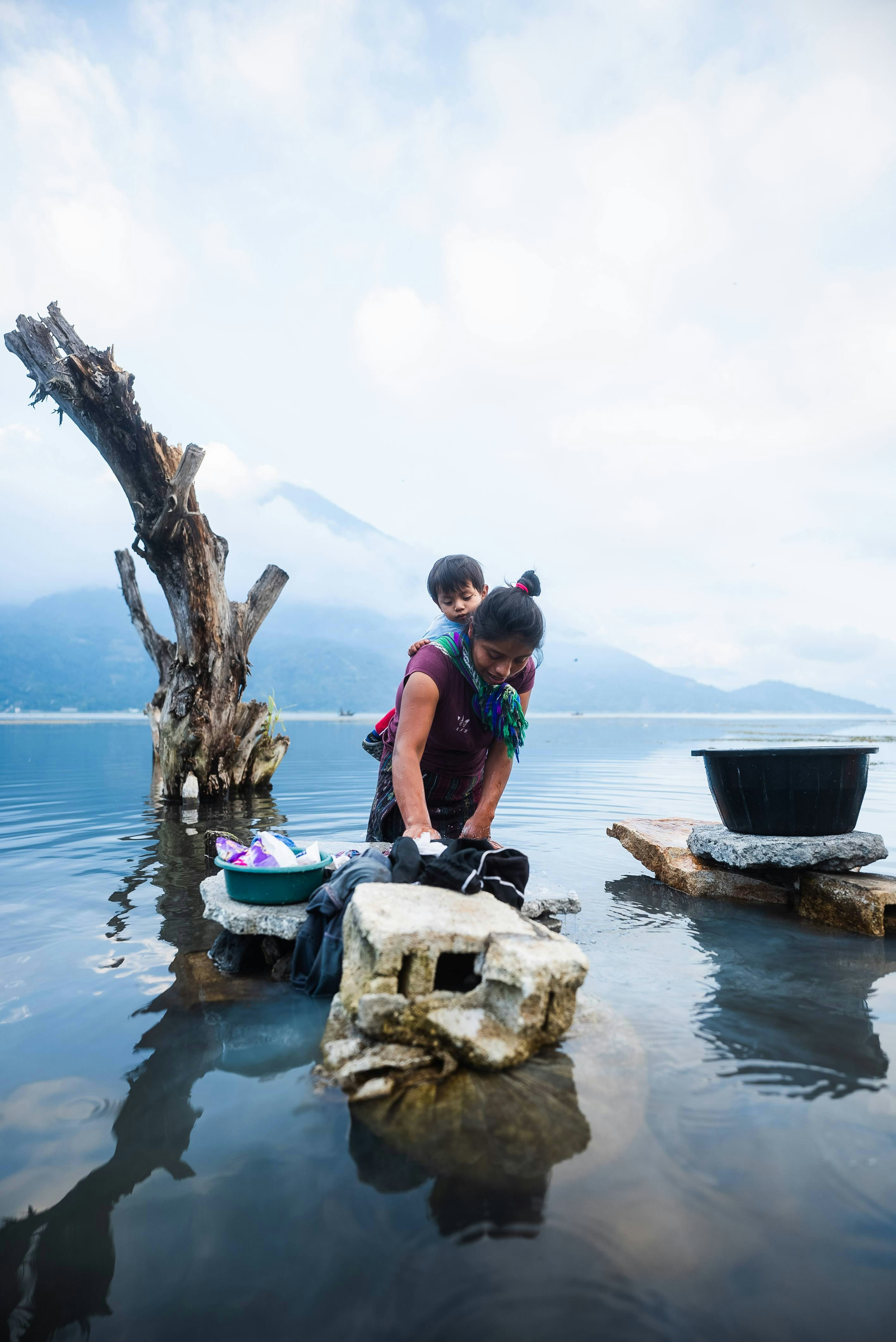 Woman washing clothes on stones in a serene lake, with a child on her back. A weathered tree stands nearby, framed by distant mountains.