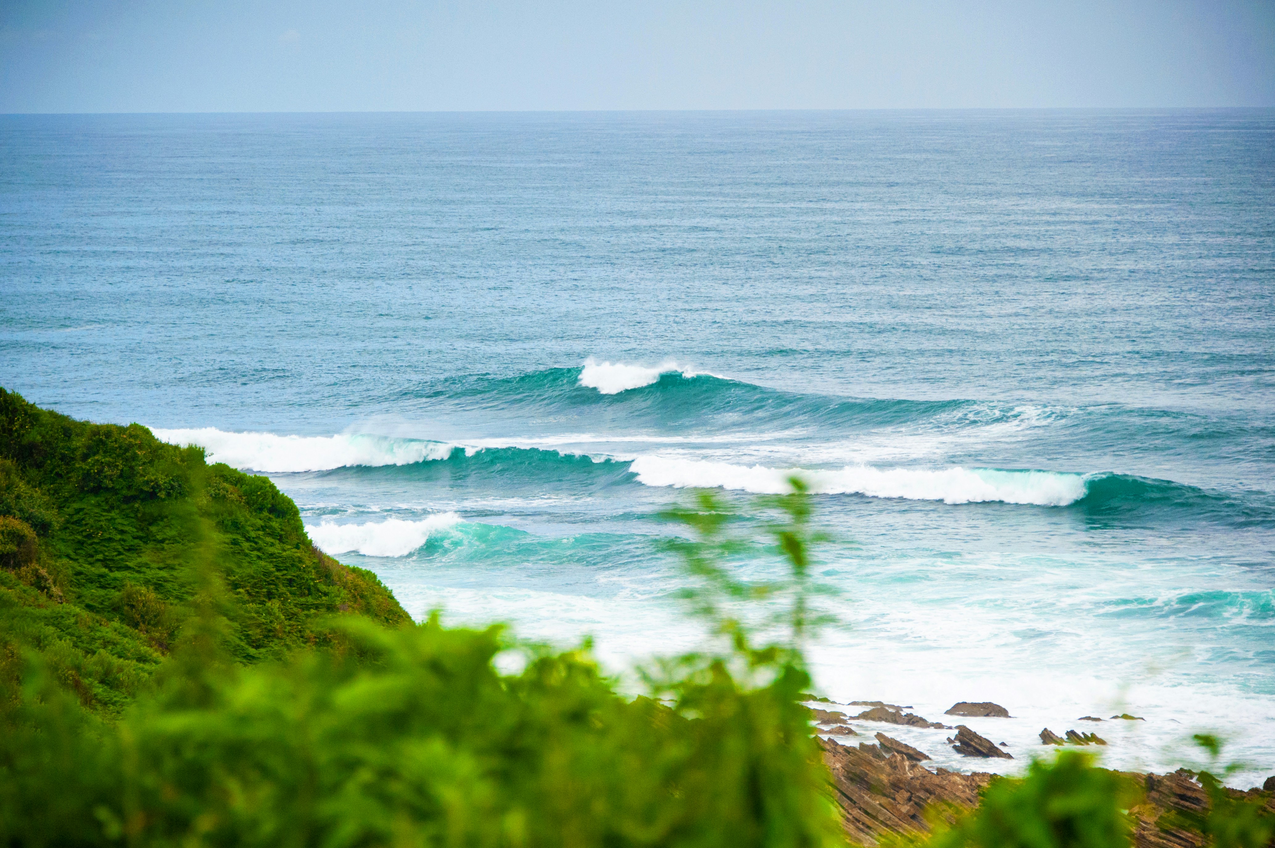 Waves gently rolling towards a rocky coastline framed by lush greenery under a soft, overcast sky.