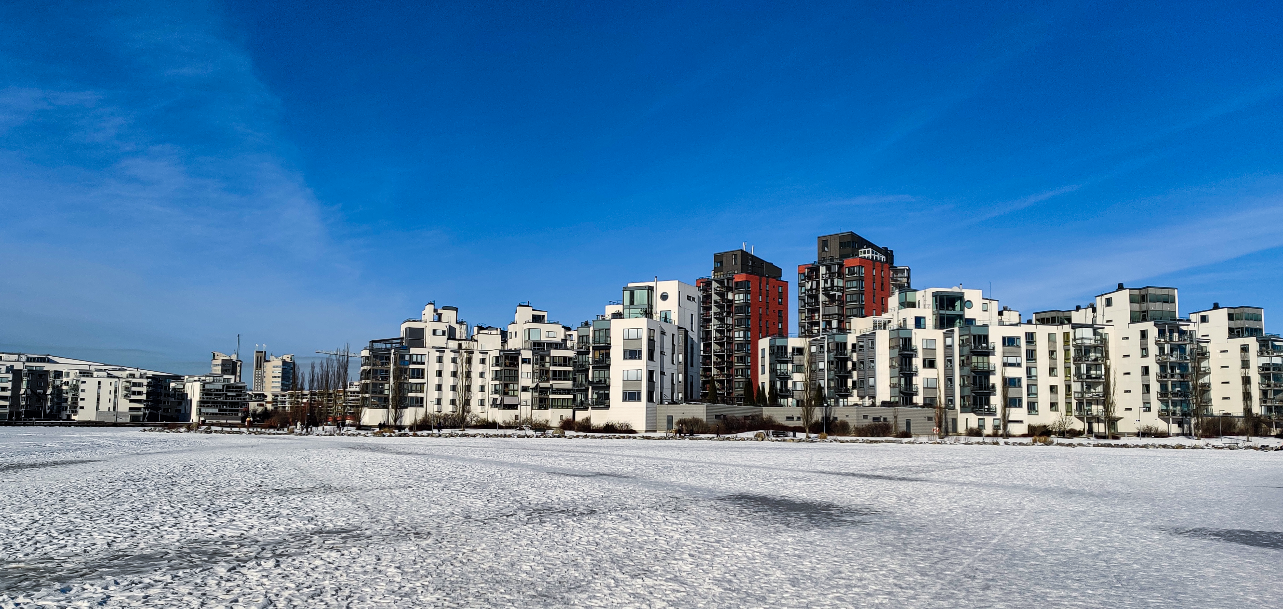 Modern apartment buildings line a snowy, open landscape under a vivid blue sky.