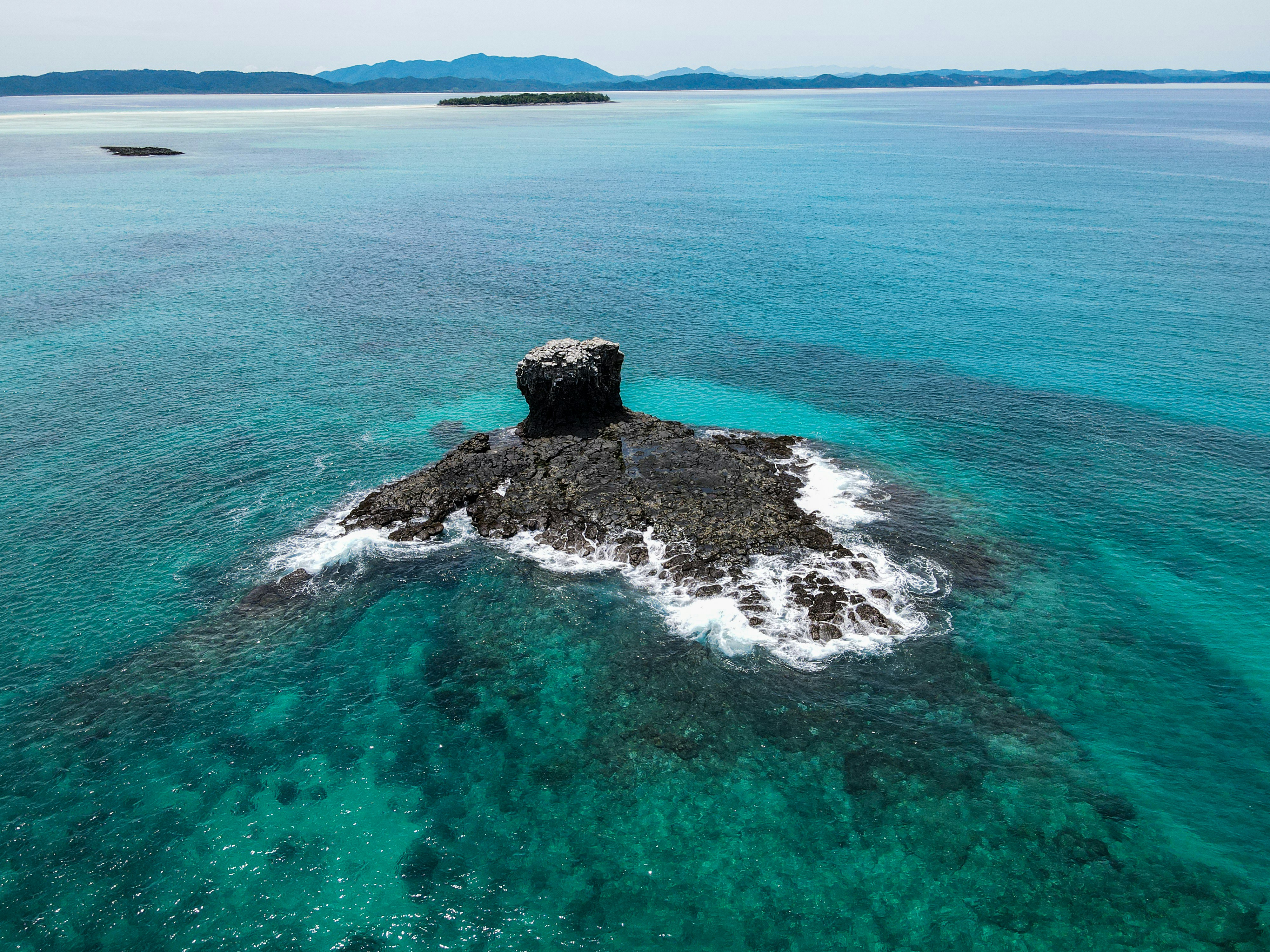 Rocky outcrop surrounded by vibrant turquoise sea and distant islands under a clear sky.