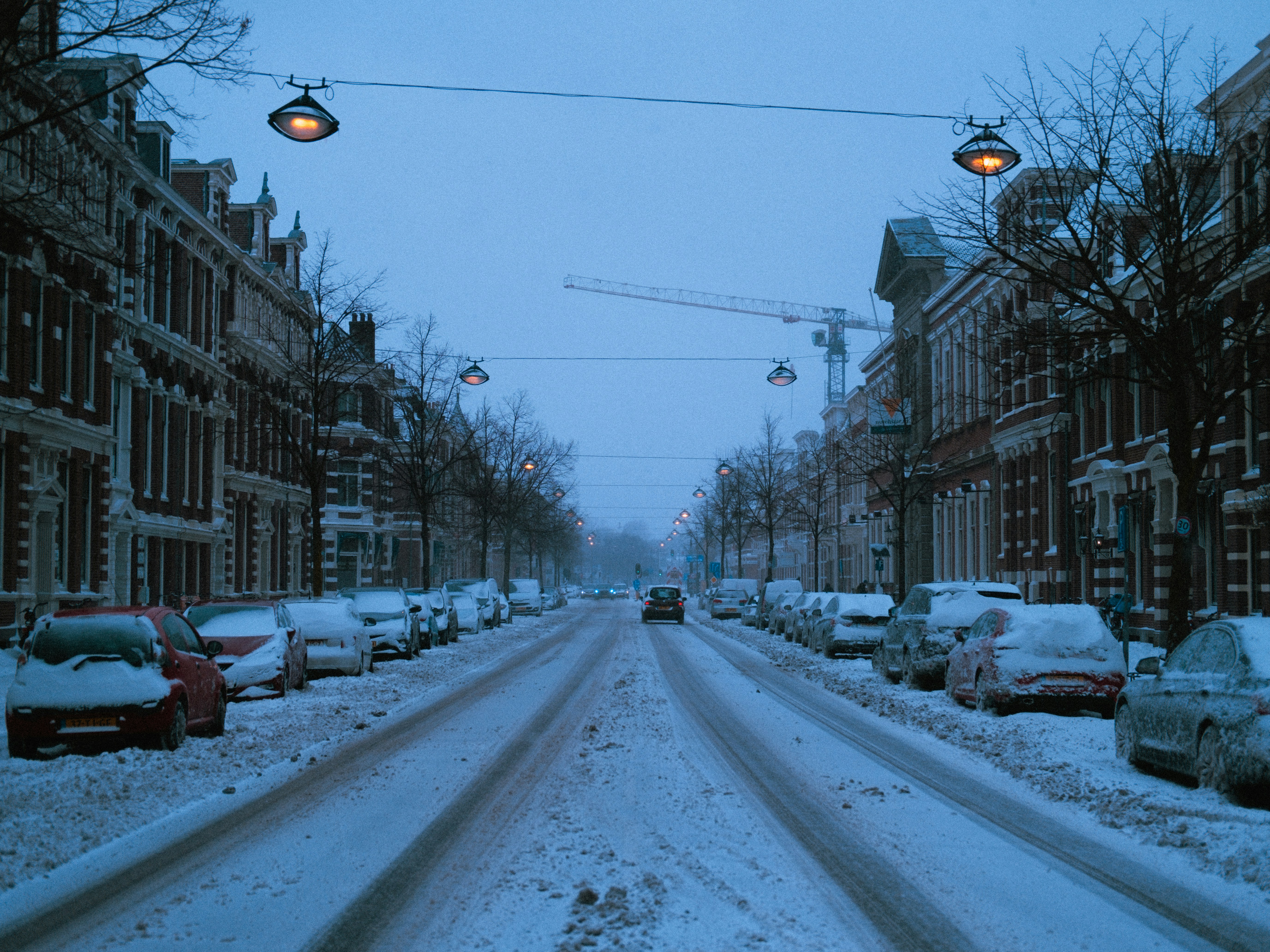 Snow-covered street lined with historic buildings and parked cars, illuminated by street lamps on a chilly evening.