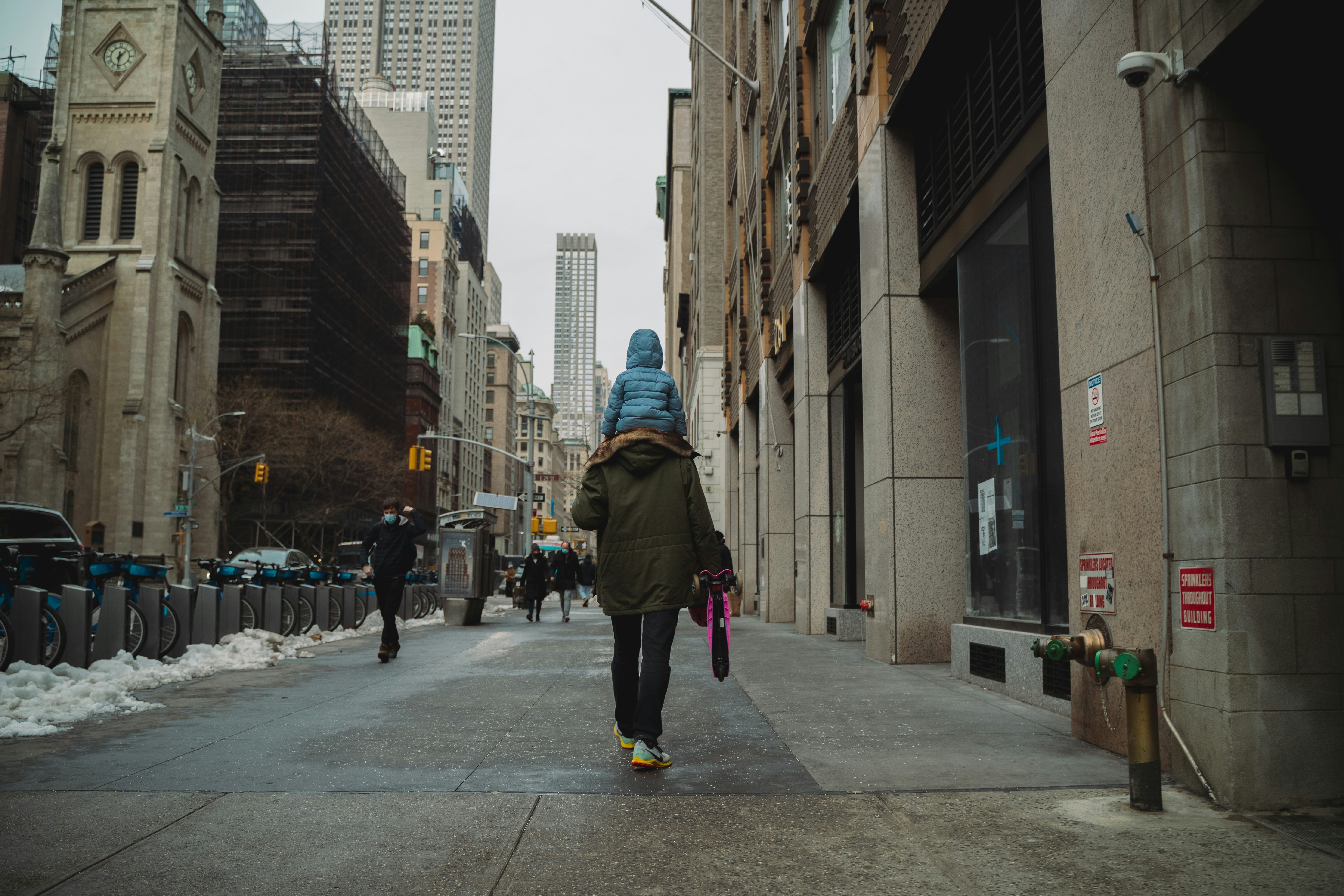 In this urban scene, a parent walks down a bustling city street with their child perched on their shoulders, creating a heartwarming focal point against the towering cityscape. The image captures the contrast between the muted tones of the buildings and the vibrant colors of the child's jacket, all under the soft, overcast lighting that adds a calming atmosphere. The composition highlights the blend of human connection and architectural grandeur, making it visually striking.