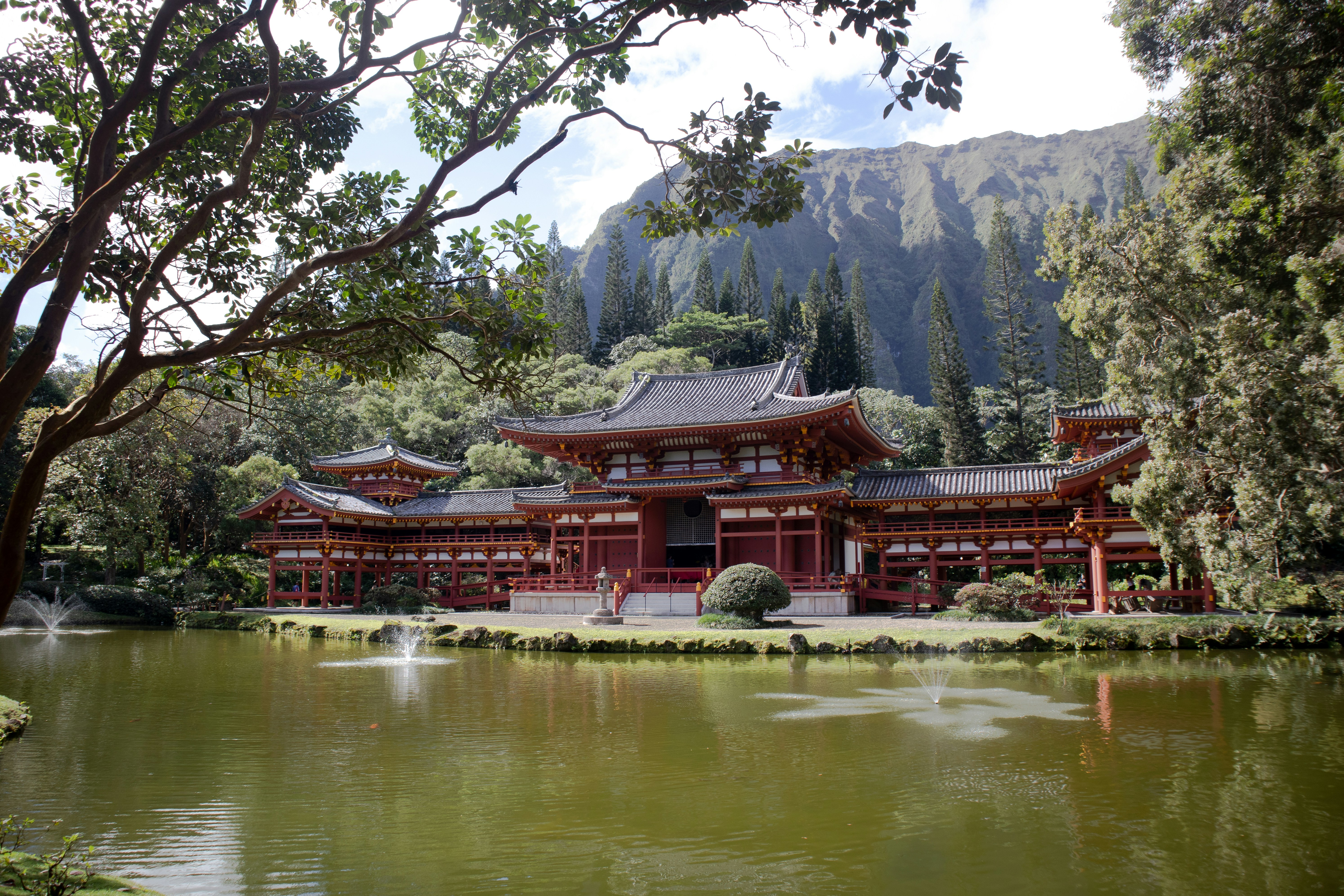 Traditional Japanese temple nestled beside a tranquil pond, surrounded by lush greenery and majestic mountains. Water features add serenity to the landscape.