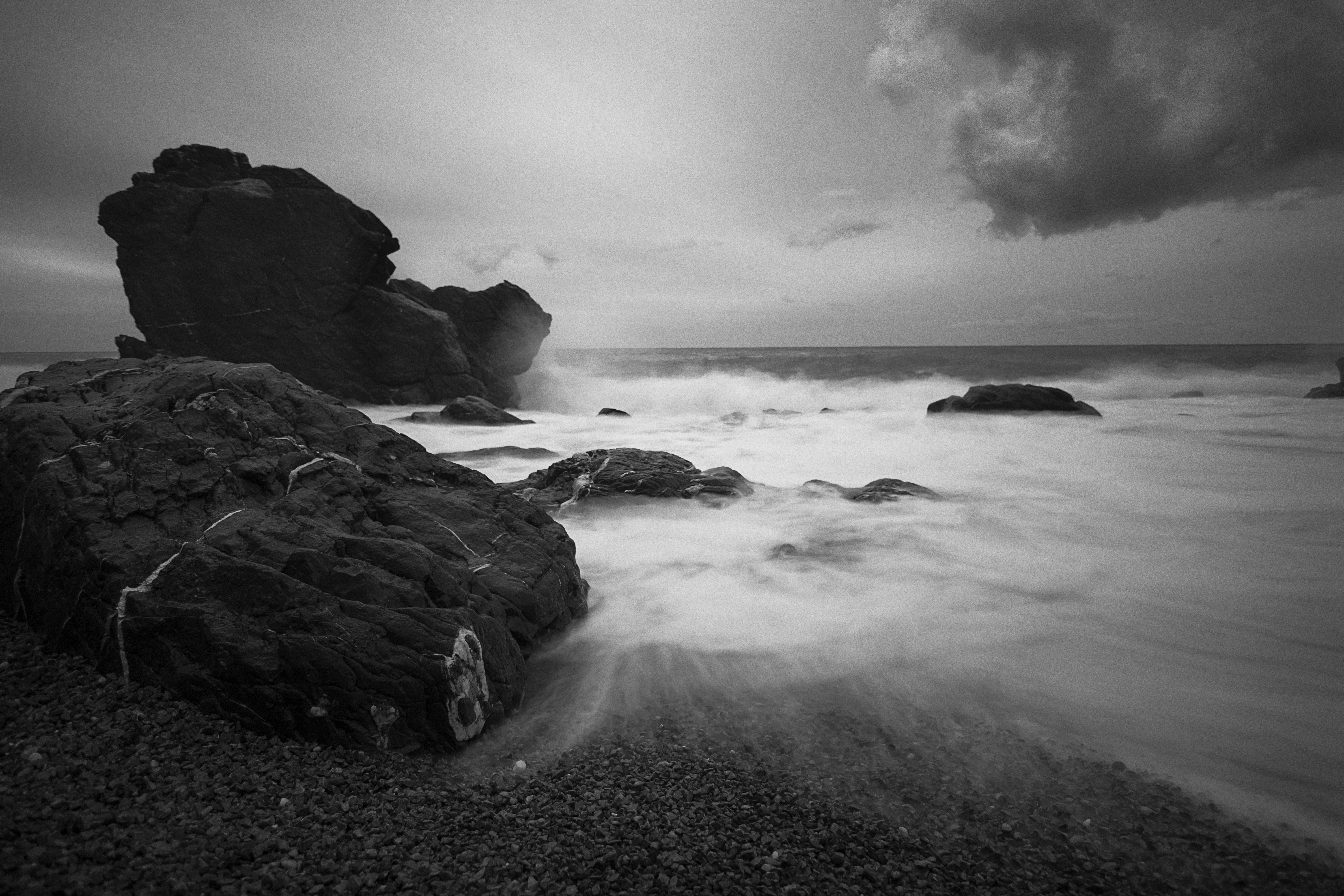 Black and white seascape with waves crashing against large coastal rocks under a cloudy sky.