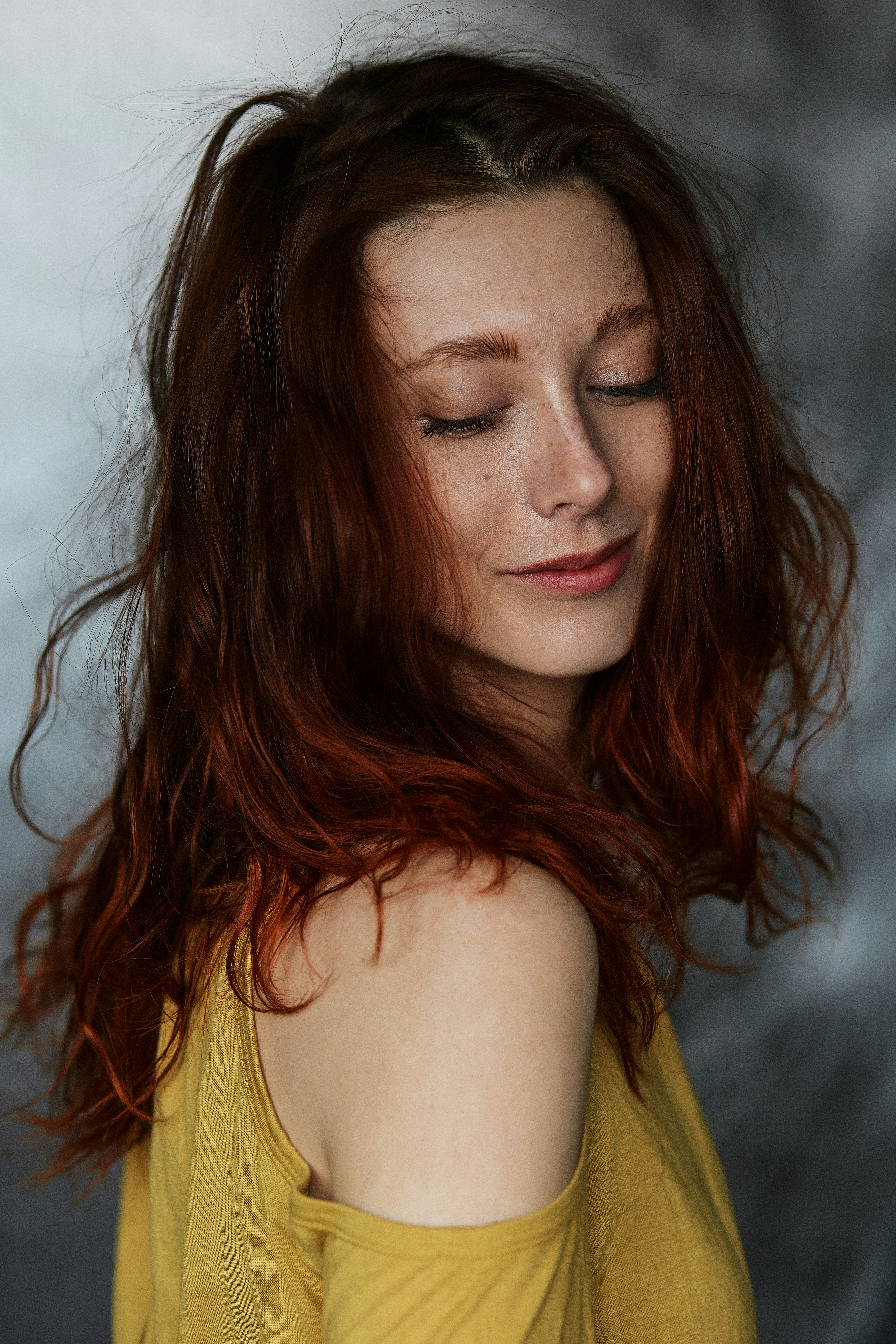 Young woman with flowing red hair and freckles gazes softly to the side, wearing an off-shoulder yellow top against a blurred backdrop.