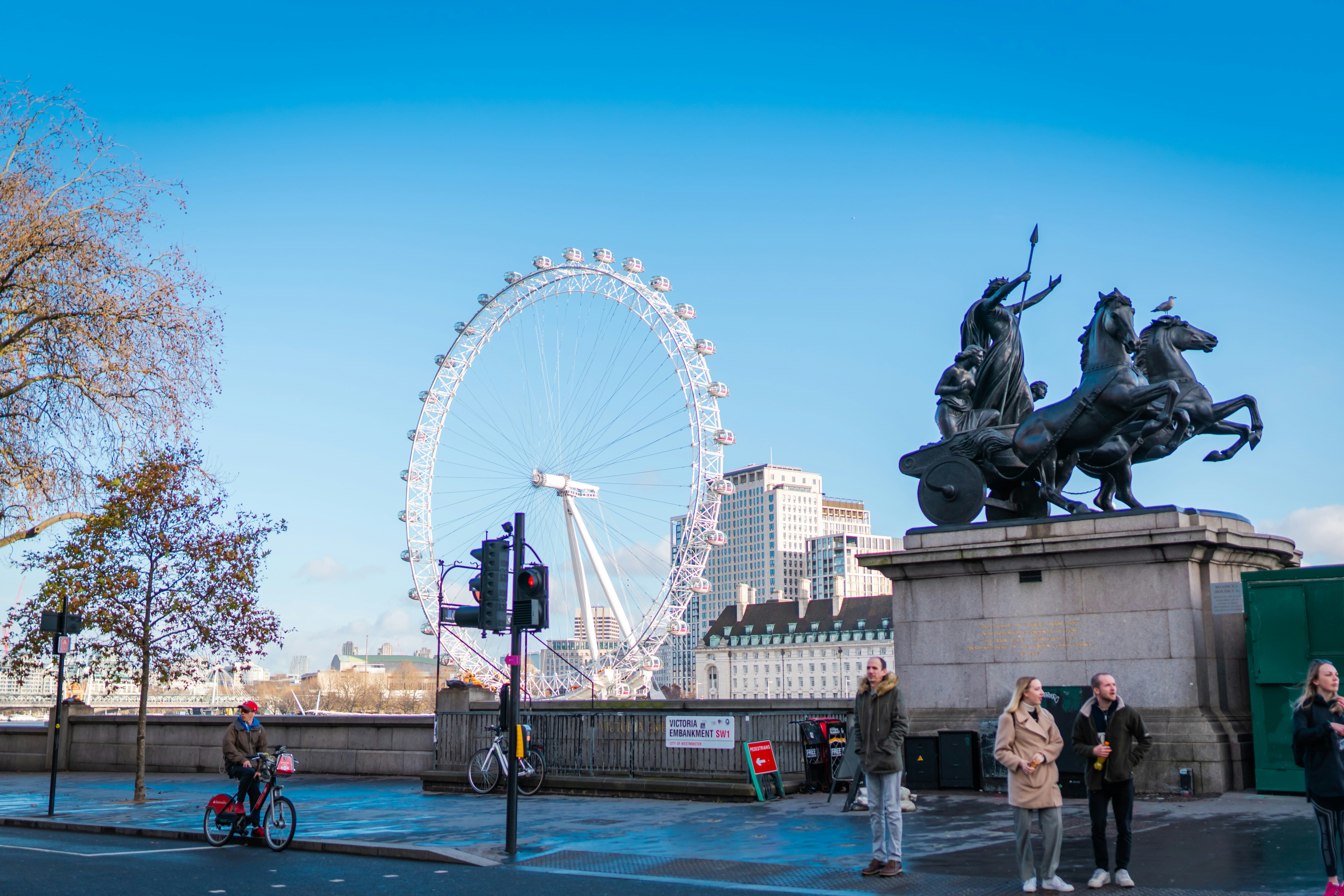 A historic statue of a chariot and horses stands prominently in the foreground, while the London Eye looms in the background against a clear blue sky.