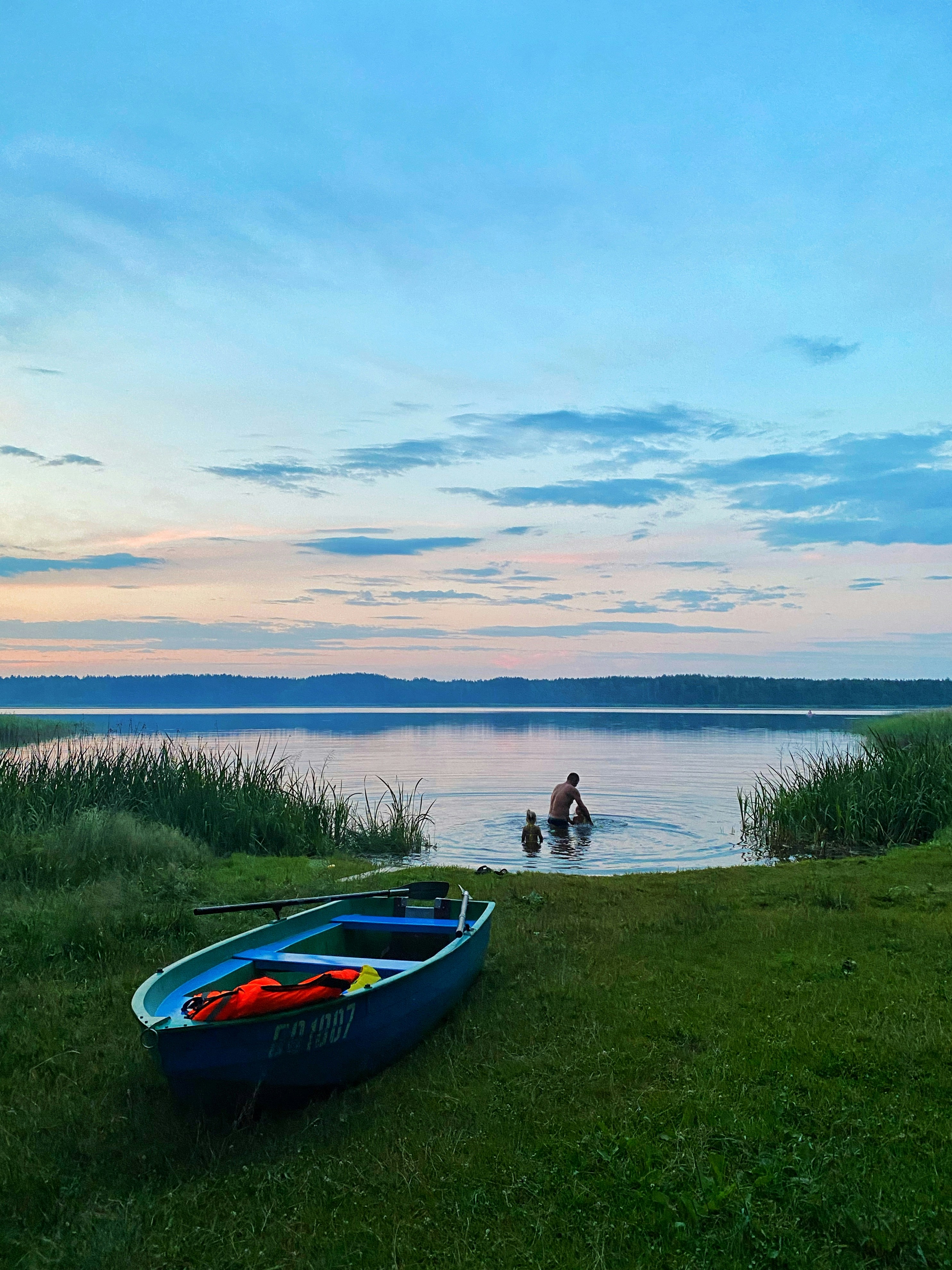 A tranquil lakeside scene featuring a man and a dog enjoying the water, with a small blue boat resting on the grass. The sky transitions from day to night, creating a peaceful atmosphere.