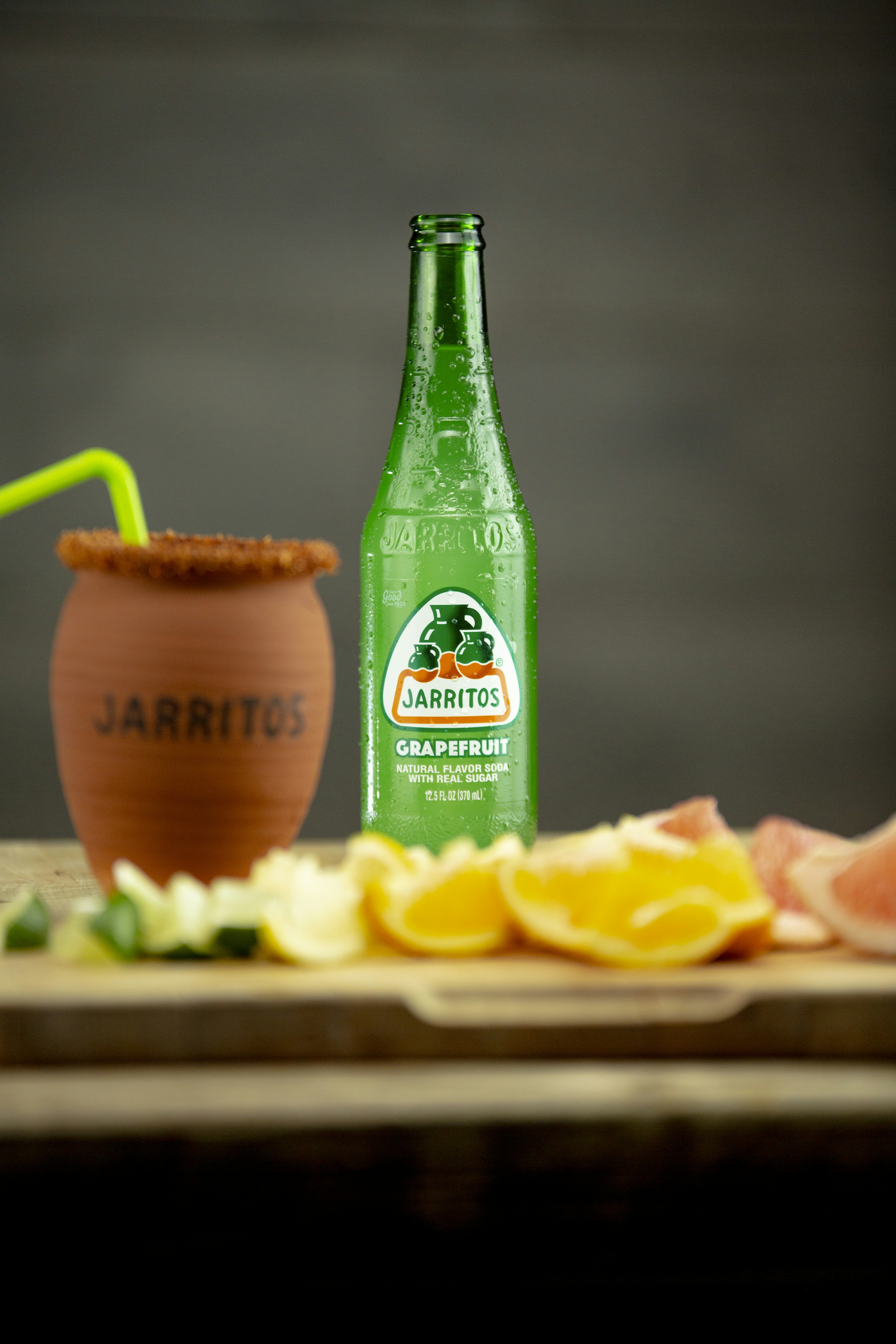 A refreshing Jarritos grapefruit soda bottle stands prominently next to a clay cup adorned with a rim of chili powder and a colorful array of citrus fruits. 