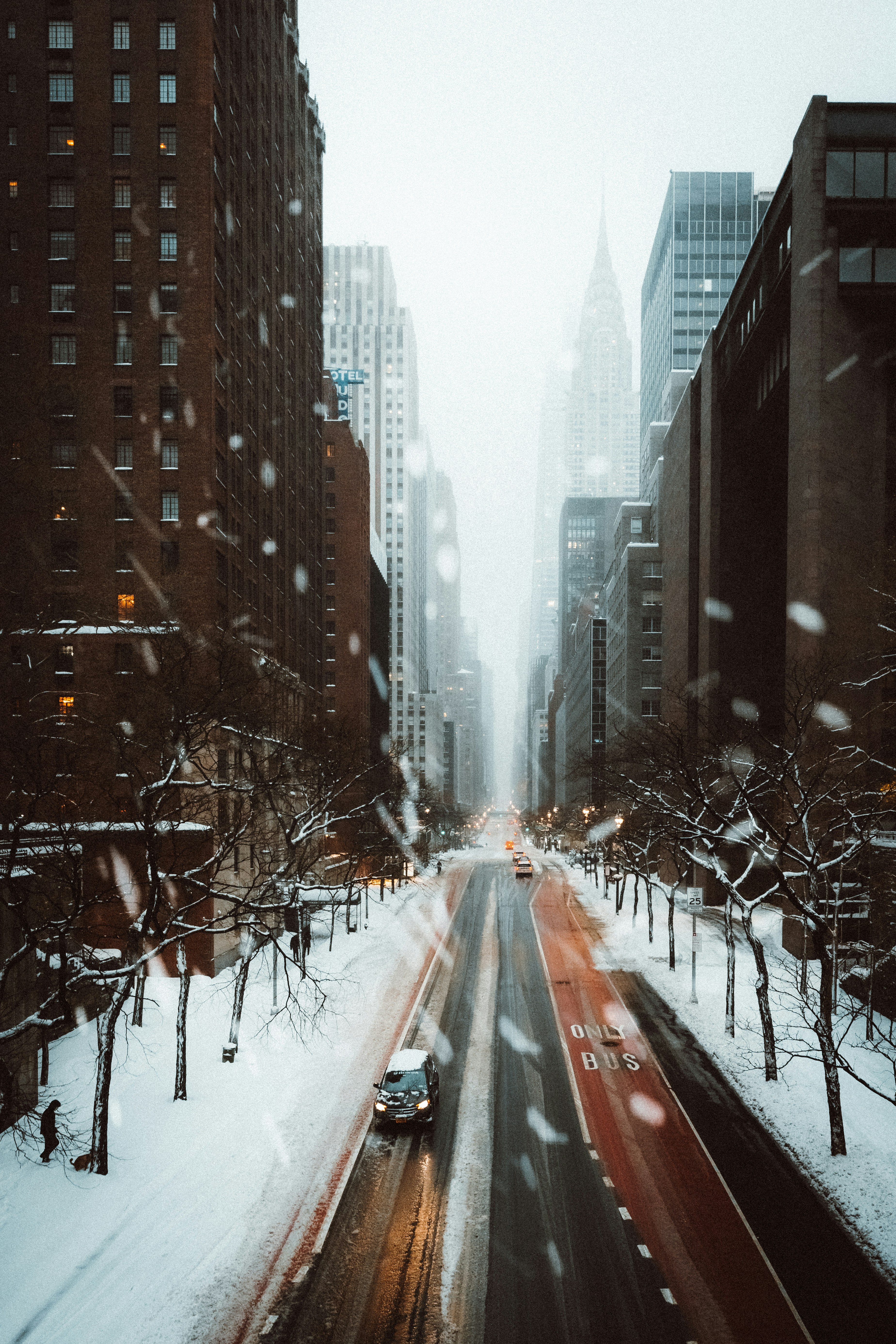 Snow-covered city street with blurred vehicles navigating through a winter storm, framed by towering skyscrapers. The iconic Empire State Building looms in the background.