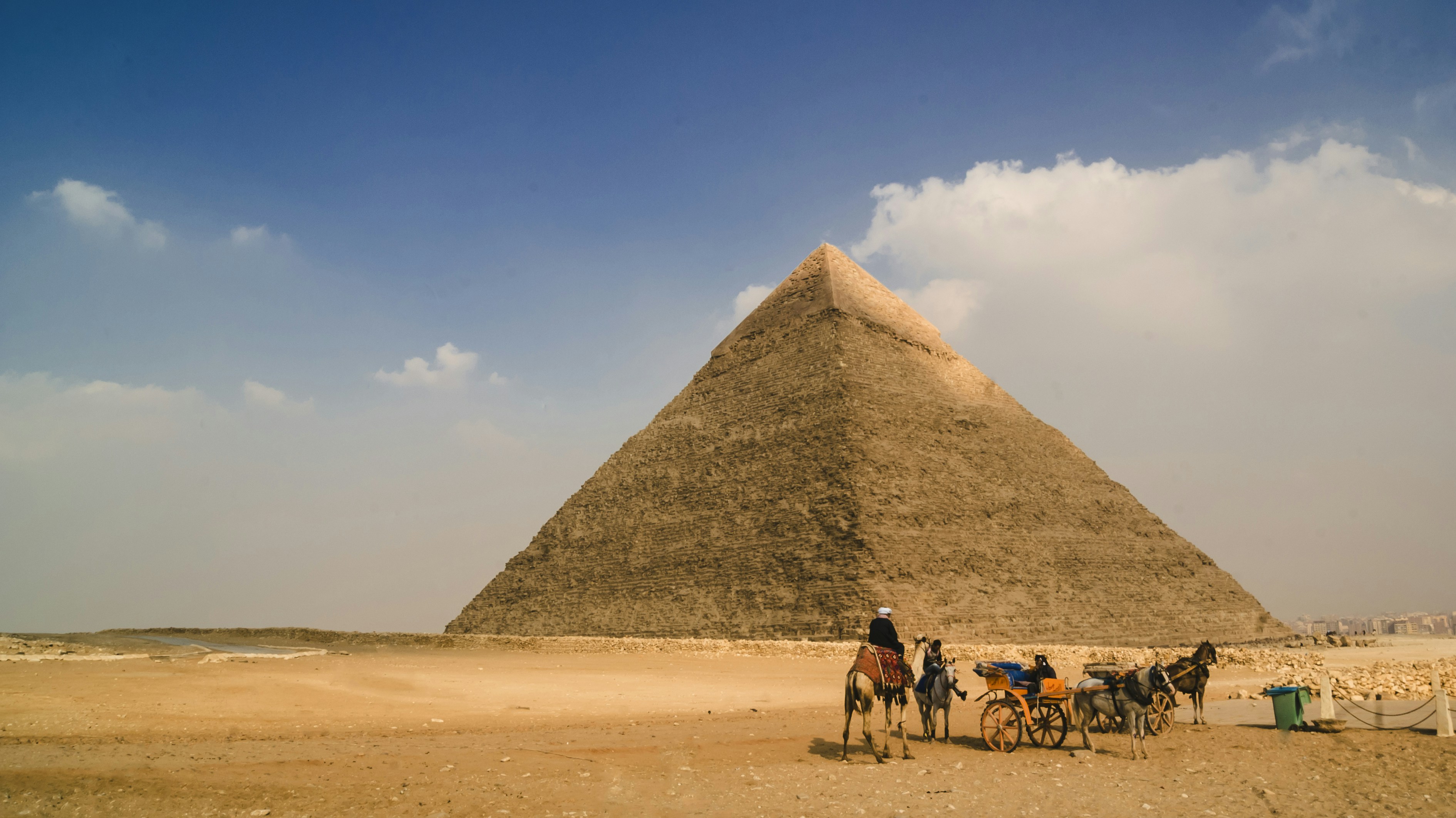 Riders on horseback traverse sandy terrain near a majestic pyramid under a clear blue sky.