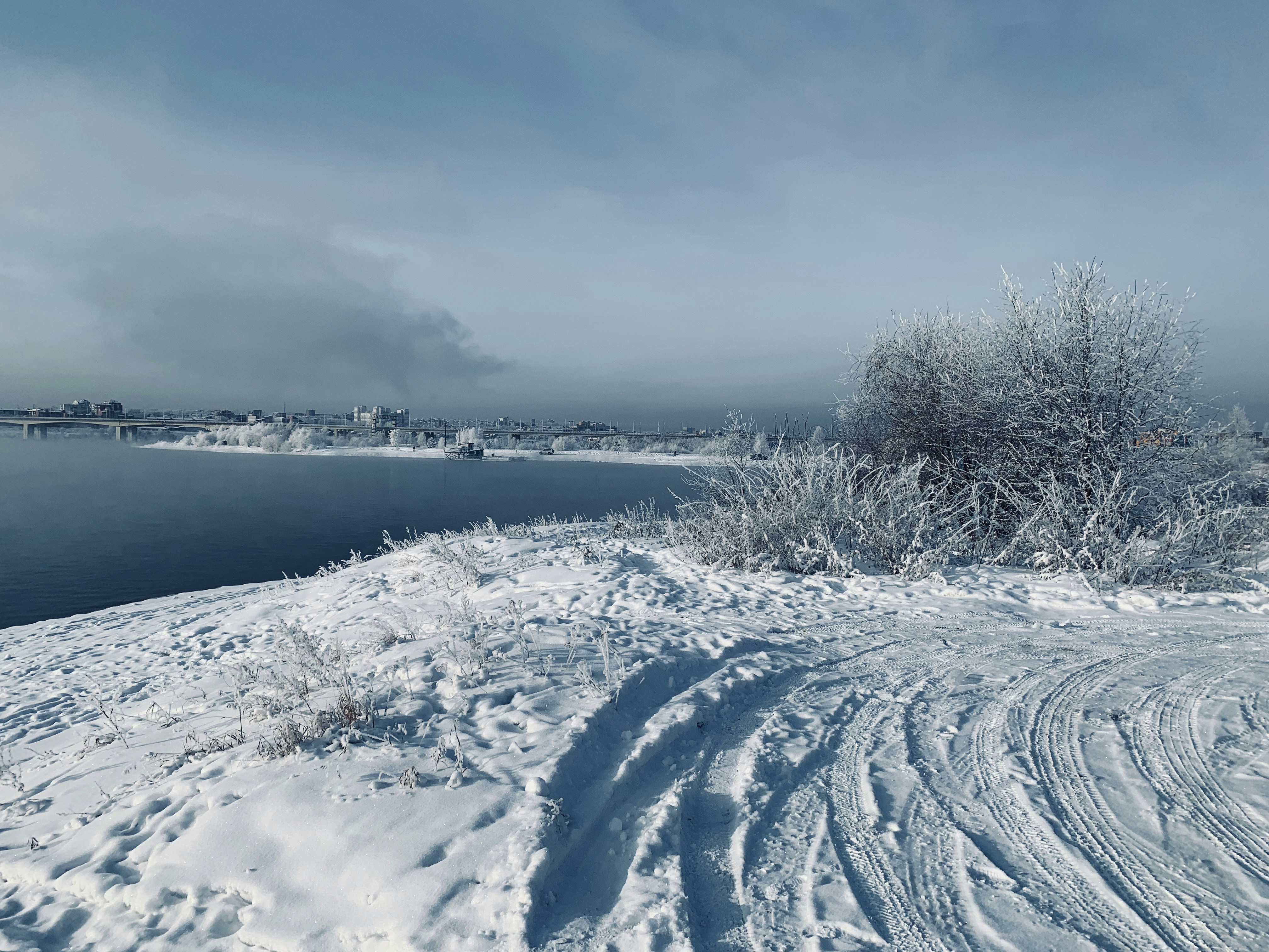 Snow-covered landscape with riverbank and frosted trees under a clear blue sky.