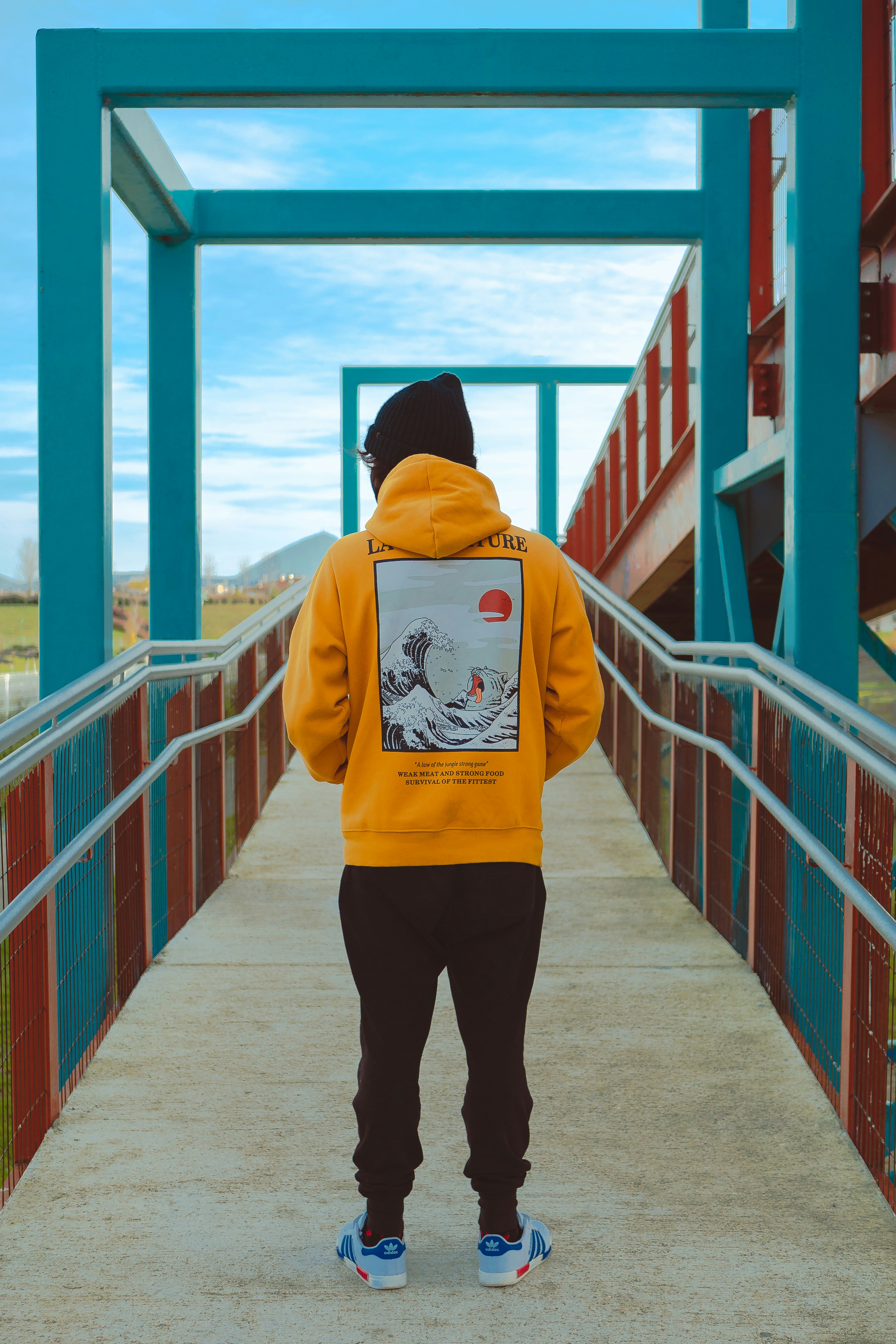 Individual in a yellow hoodie with a graphic design stands on a colorful walkway, framed by vibrant structures. The scene conveys a sense of adventure and personal style.