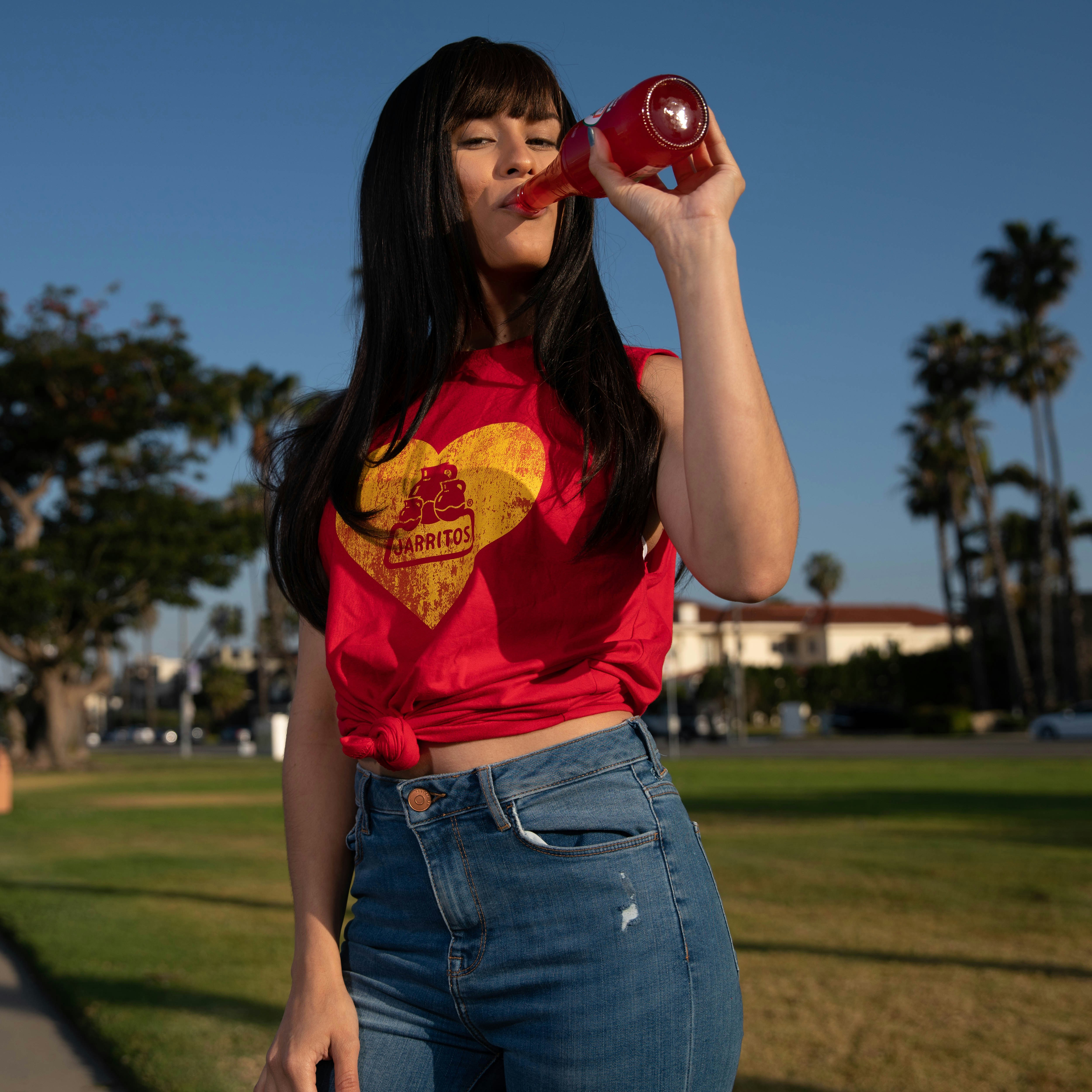 Young woman enjoying a drink while wearing a vibrant red shirt, set against a backdrop of palm trees and urban scenery.