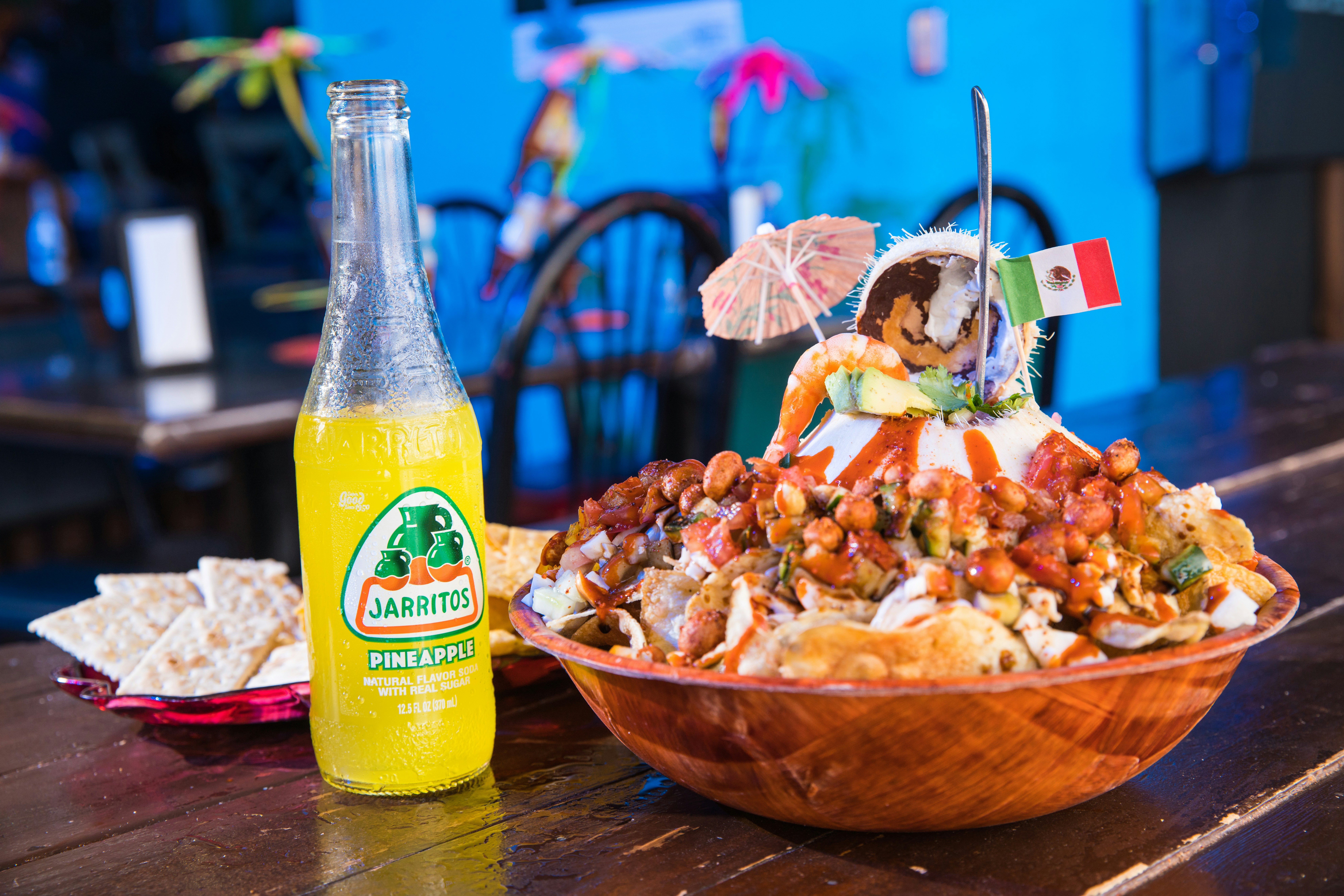 A vibrant bowl of nachos topped with beans, sauces, and garnishes, accompanied by a Jarritos pineapple soda and tortilla chips. The colorful setting enhances the festive atmosphere.