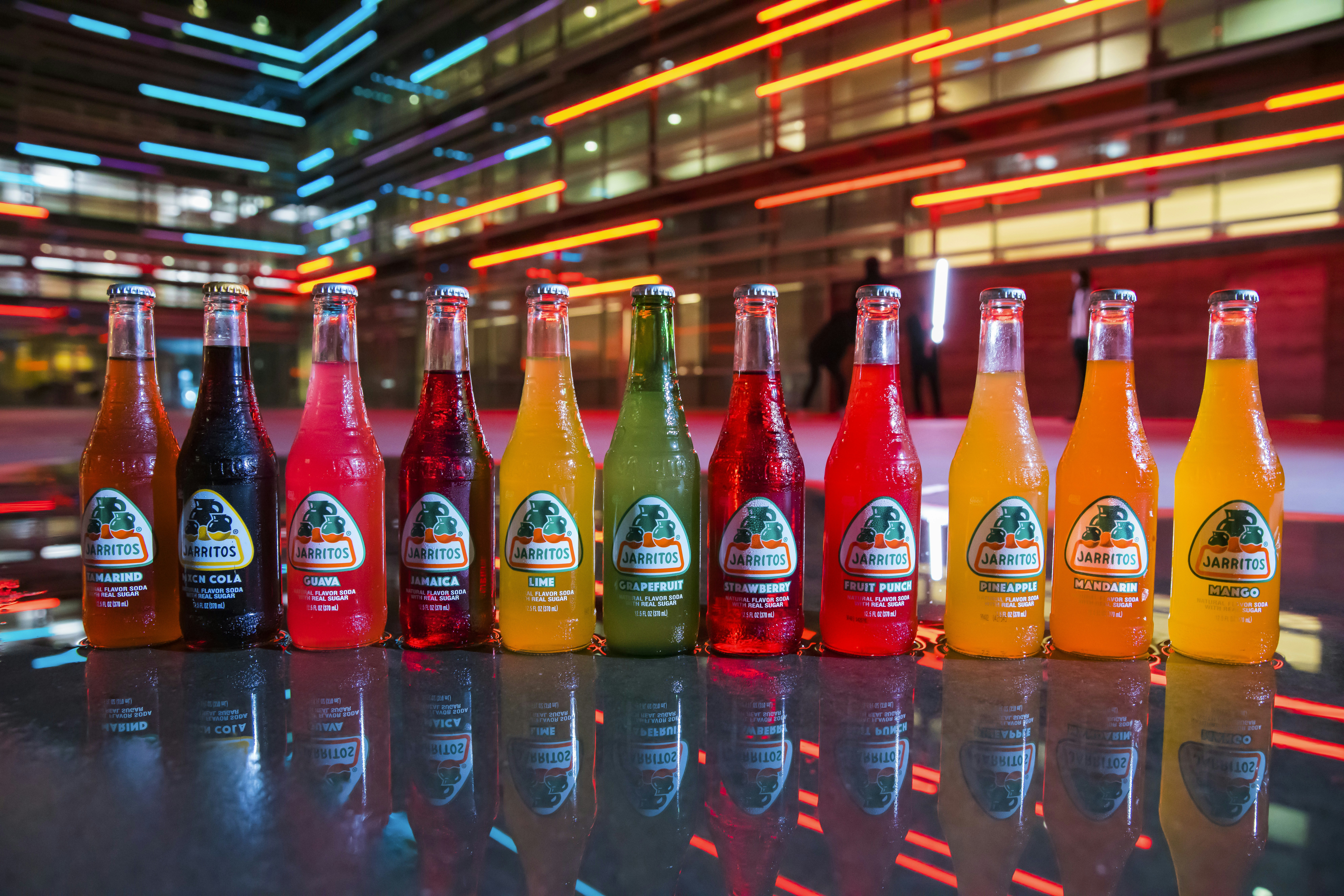 Colorful assortment of bottled beverages arranged on a reflective surface, illuminated by vibrant neon lights in the background.