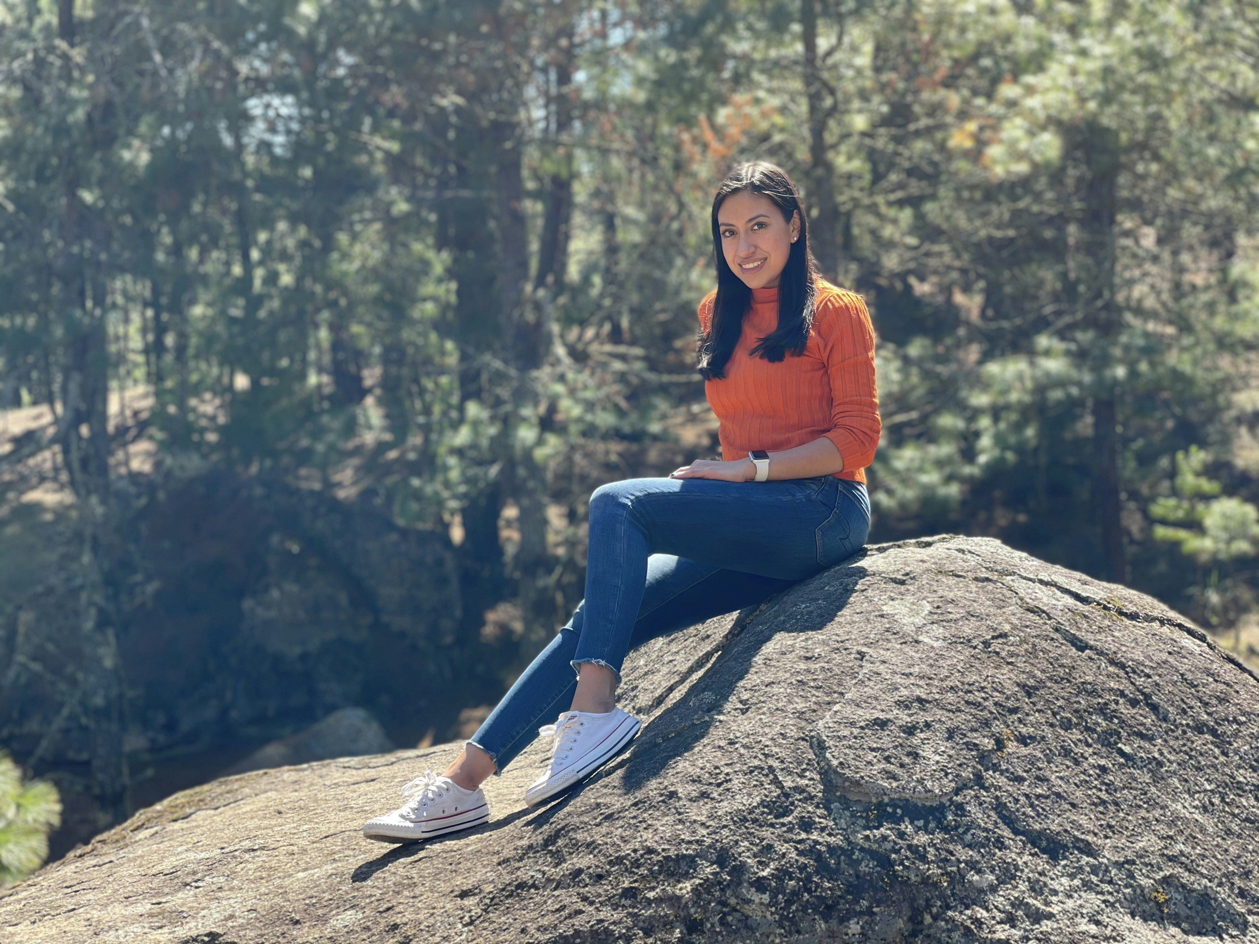 Woman in orange shirt and blue jeans sitting on a large rock in a sunlit forest.