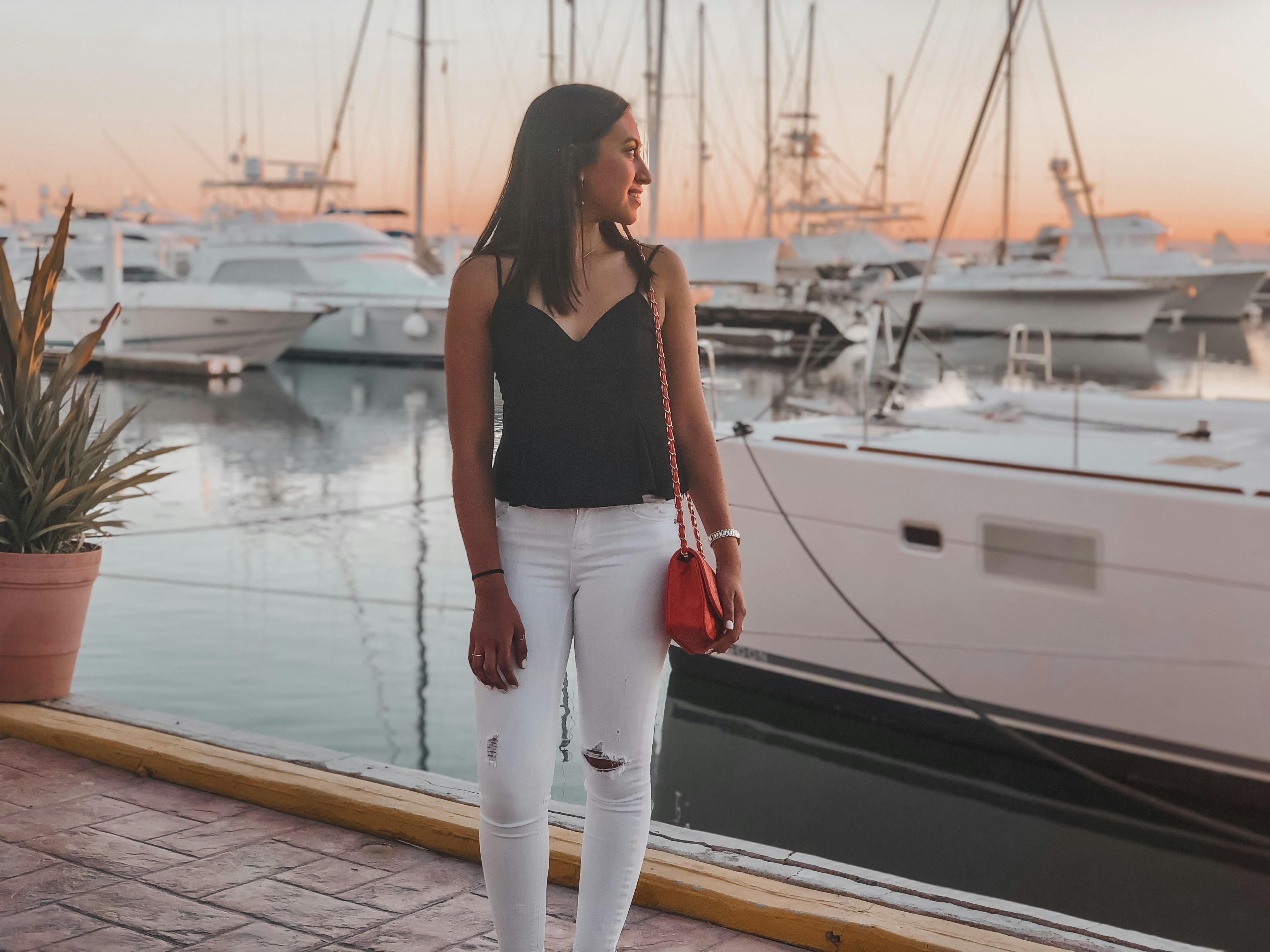 Woman in white jeans and black top stands near docked yachts at sunset.