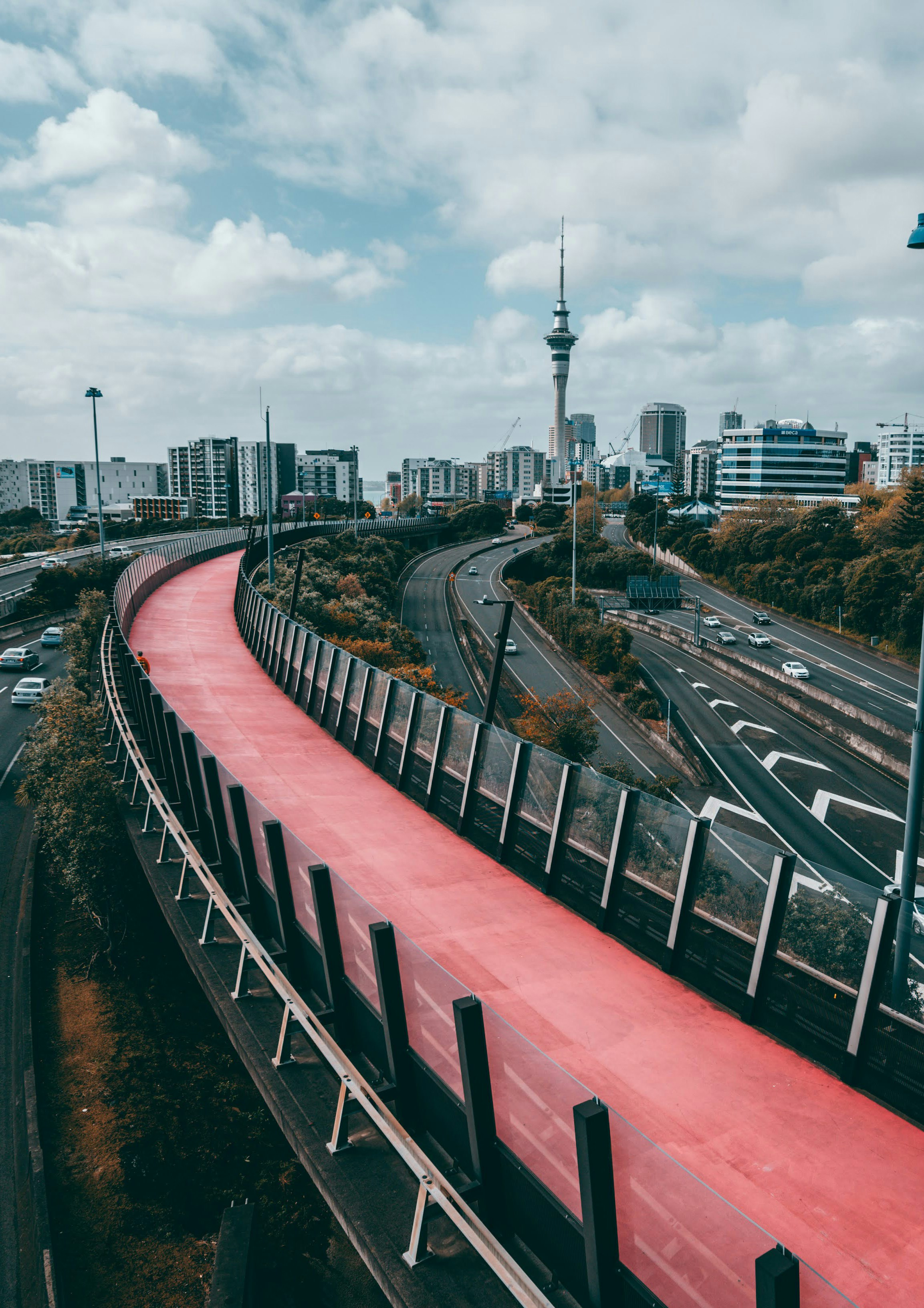 Urban landscape photograph of a red elevated walkway winding along a highway with a city skyline and a tall tower in the distance.