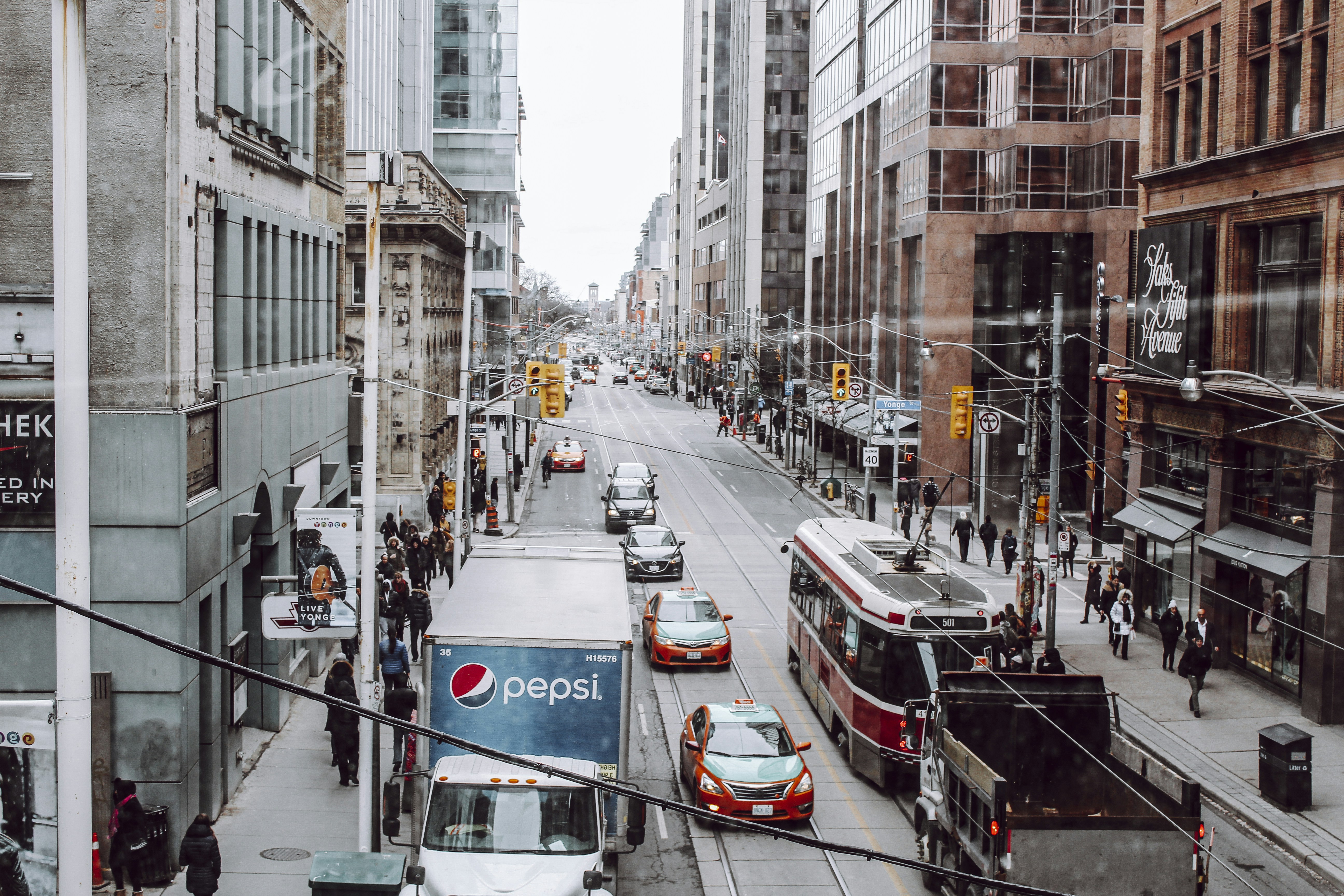 Bustling city street with streetcars, cars, and pedestrians between towering buildings on a cloudy day.