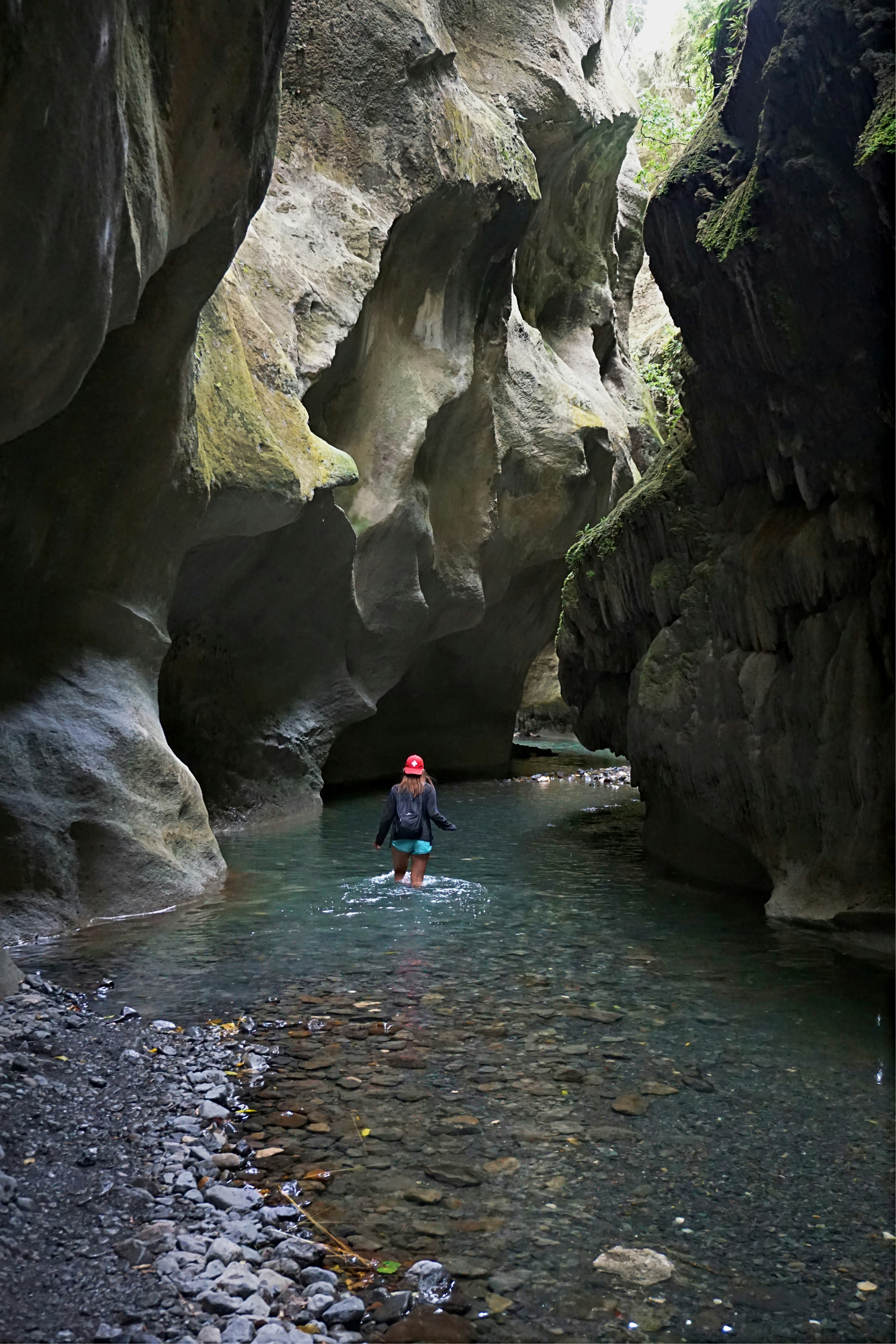 A hiker wades through a serene waterway surrounded by towering rock formations in a narrow canyon. The lush greenery adds a touch of vibrancy to the rugged landscape.